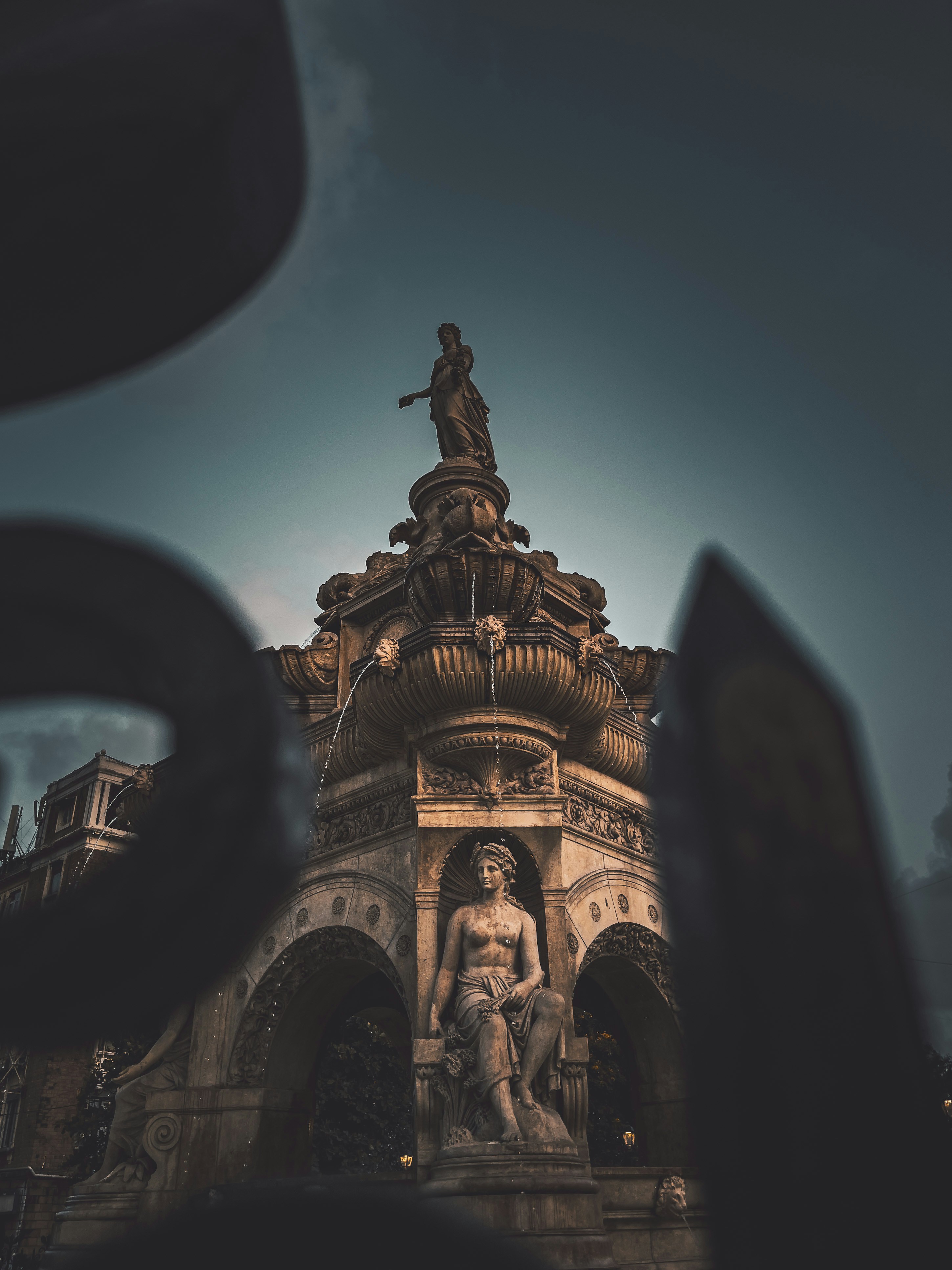 Intricate fountain featuring a central statue surrounded by ornate details, framed by a wrought iron structure. Water cascades gracefully from the top, creating a serene atmosphere.