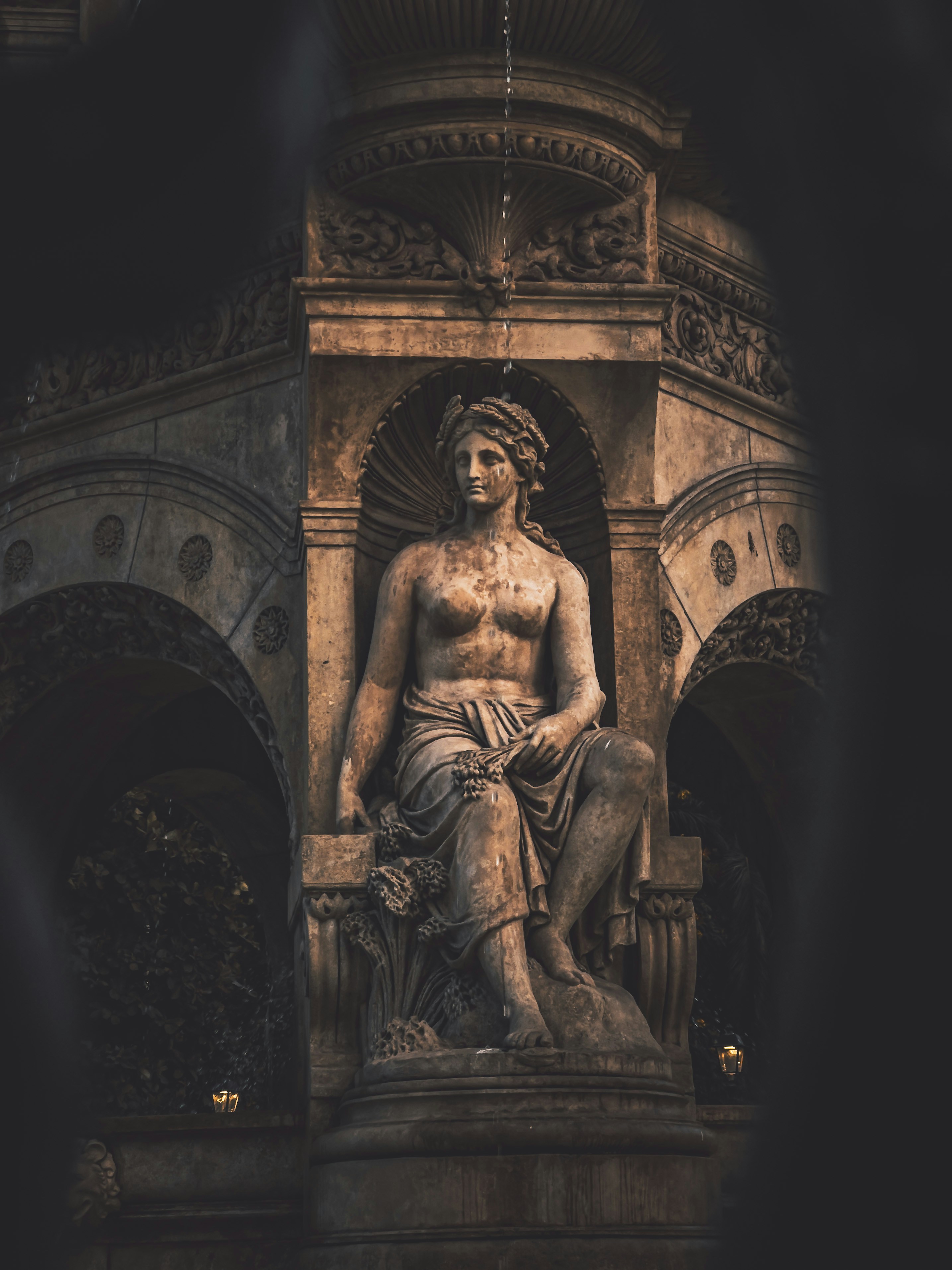 A classical statue of a seated figure adorned with floral elements, framed by ornate architecture. Water gently cascades in the background.