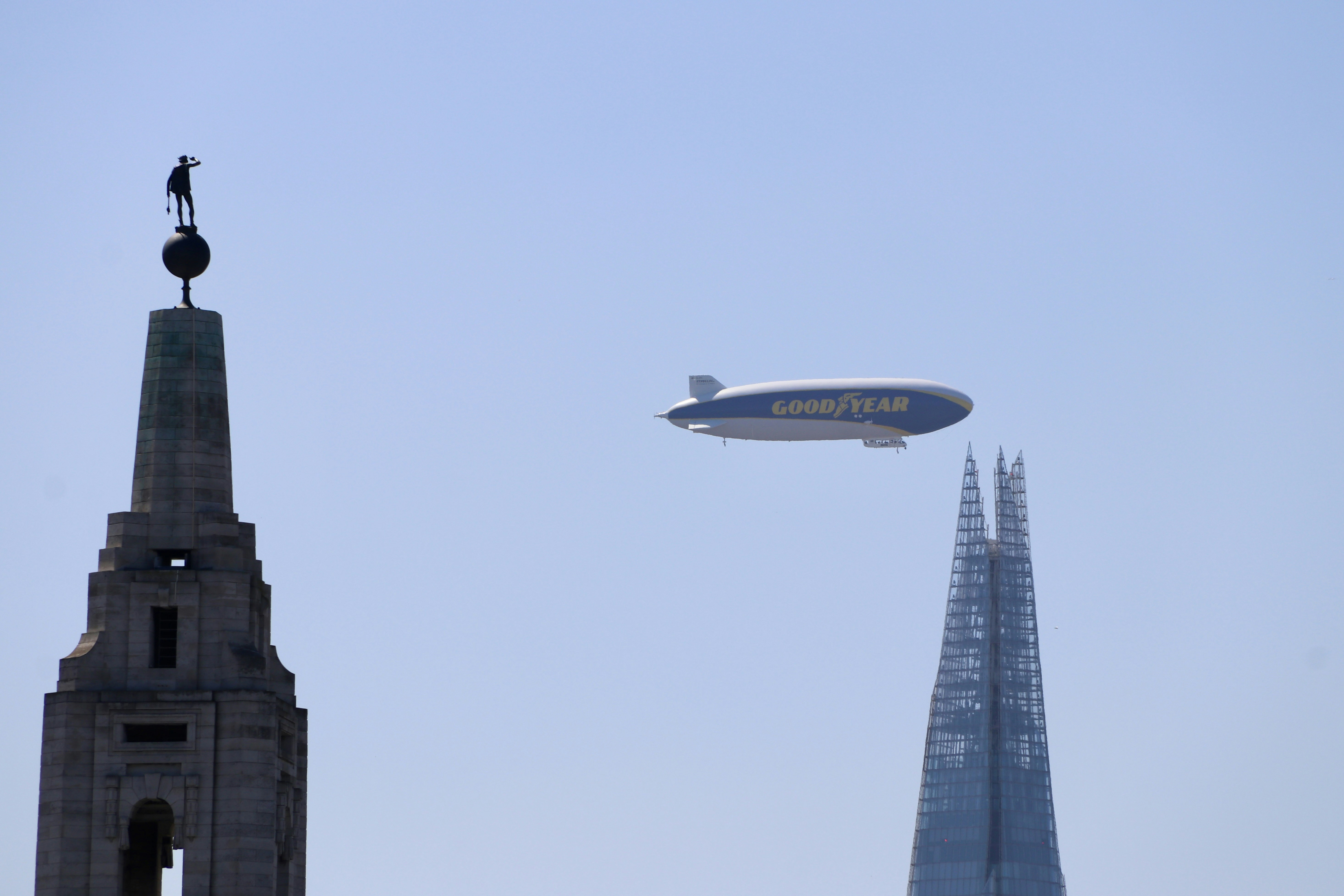 The Goodyear blimp flying over London for the British Touring Cars Championship. | an airplane flying over a tall building with a statue on top of it