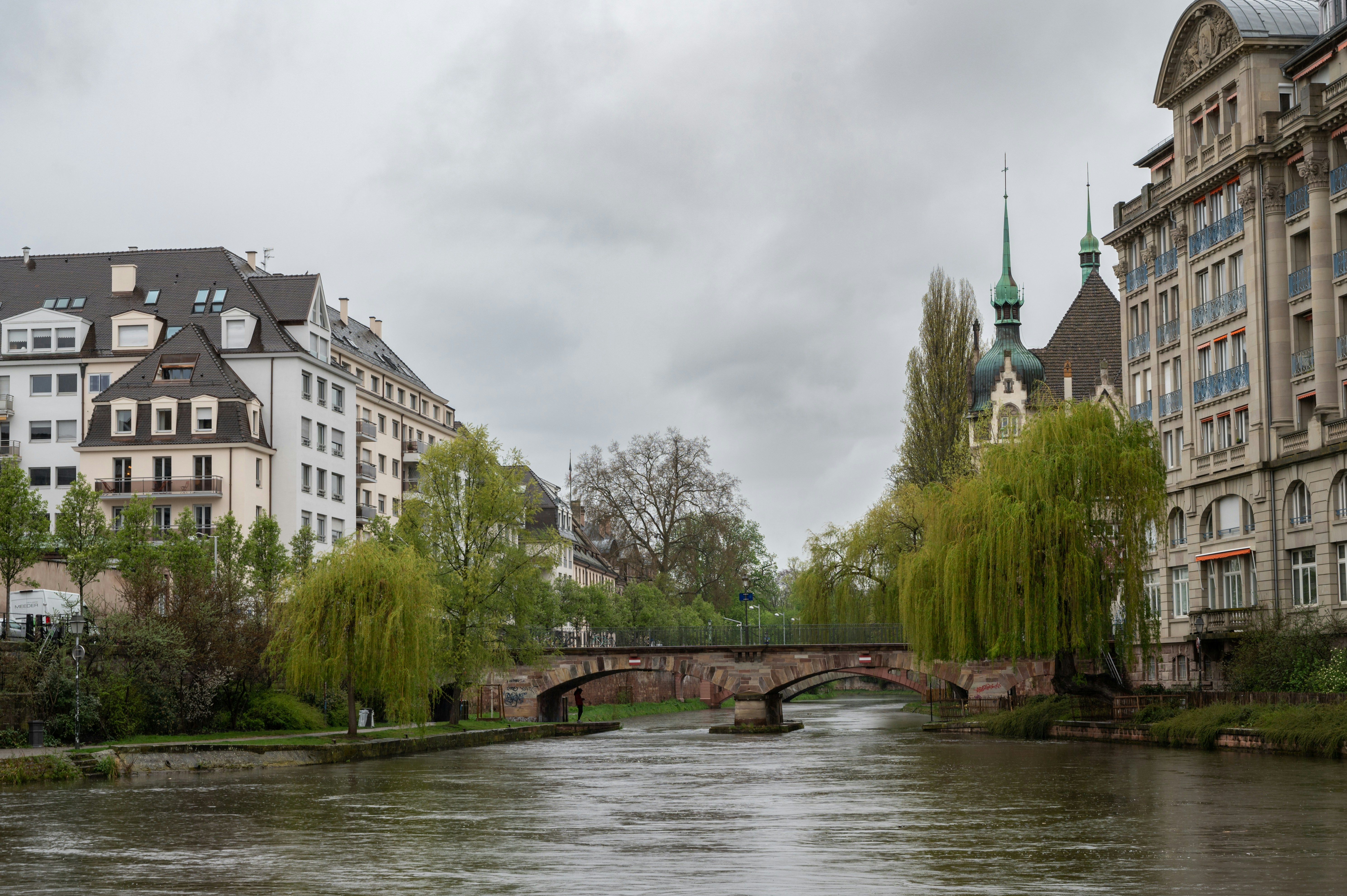 Stone bridge arches over a calm river with lush trees and historic buildings under a cloudy sky.