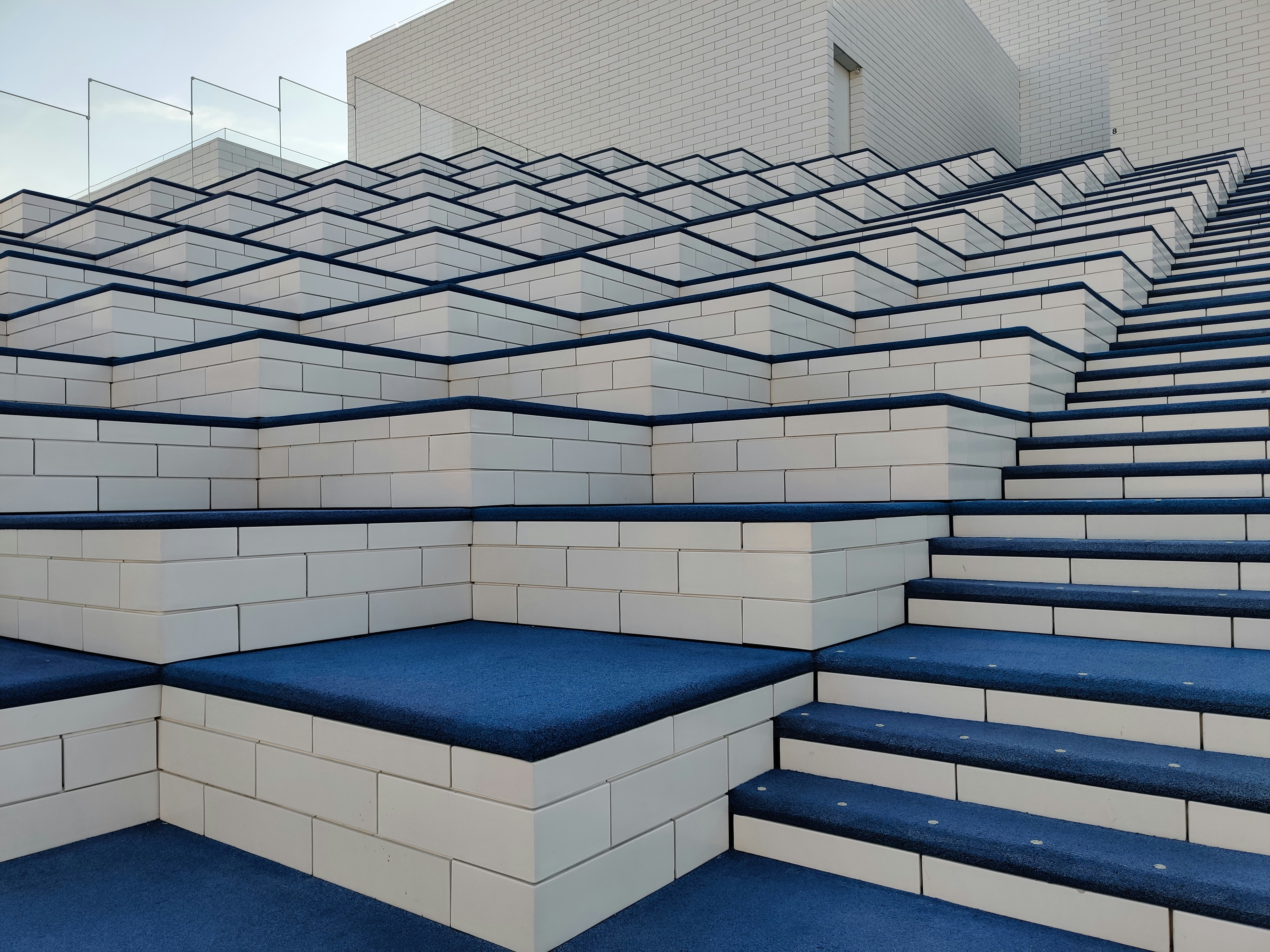 Architectural photograph of white stepped platforms with blue cushions forming a geometric, zigzag pattern that emphasizes perspective against a modern facade.