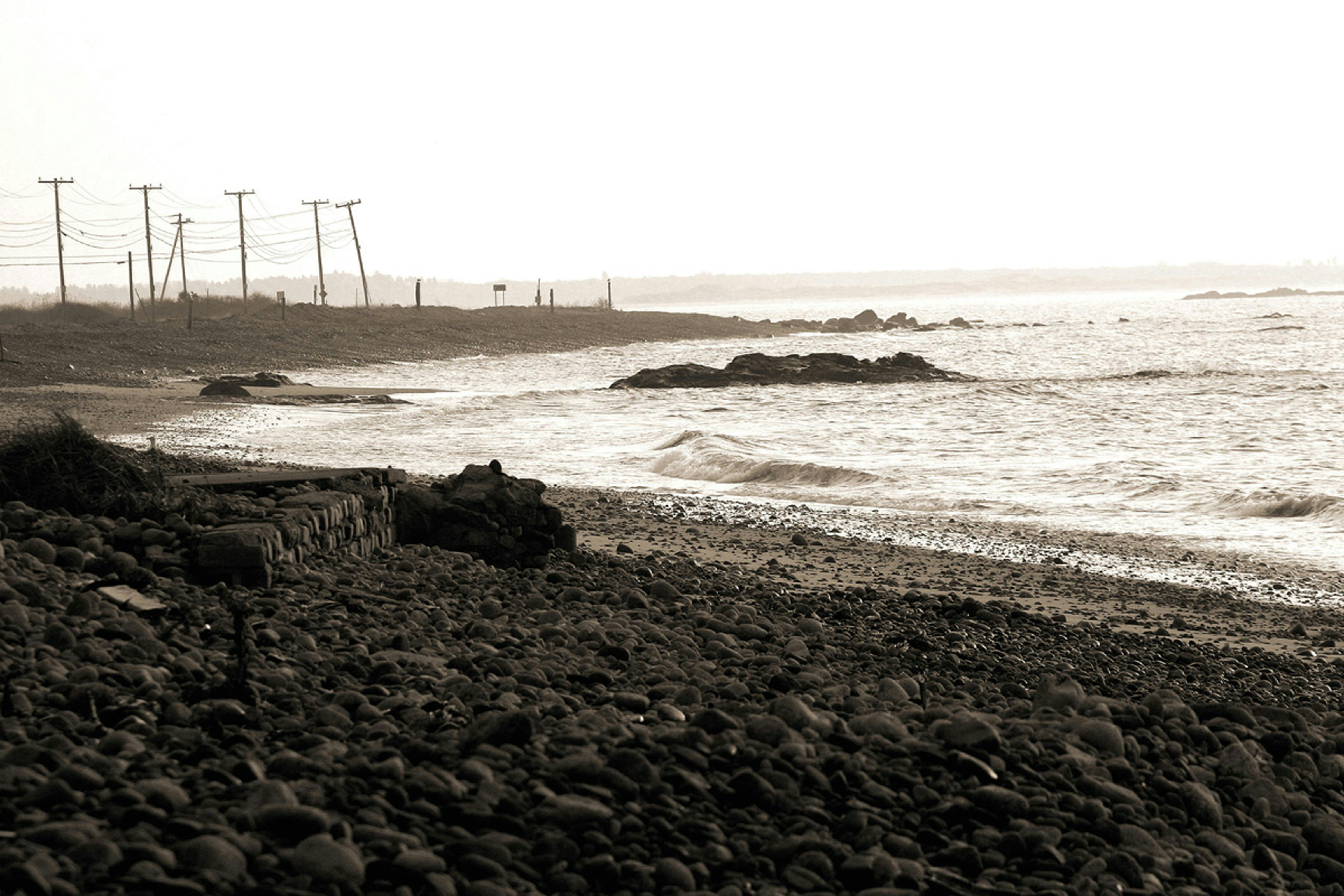 Rocky beach with gentle waves lapping at the shore, framed by distant power lines under a hazy sky.