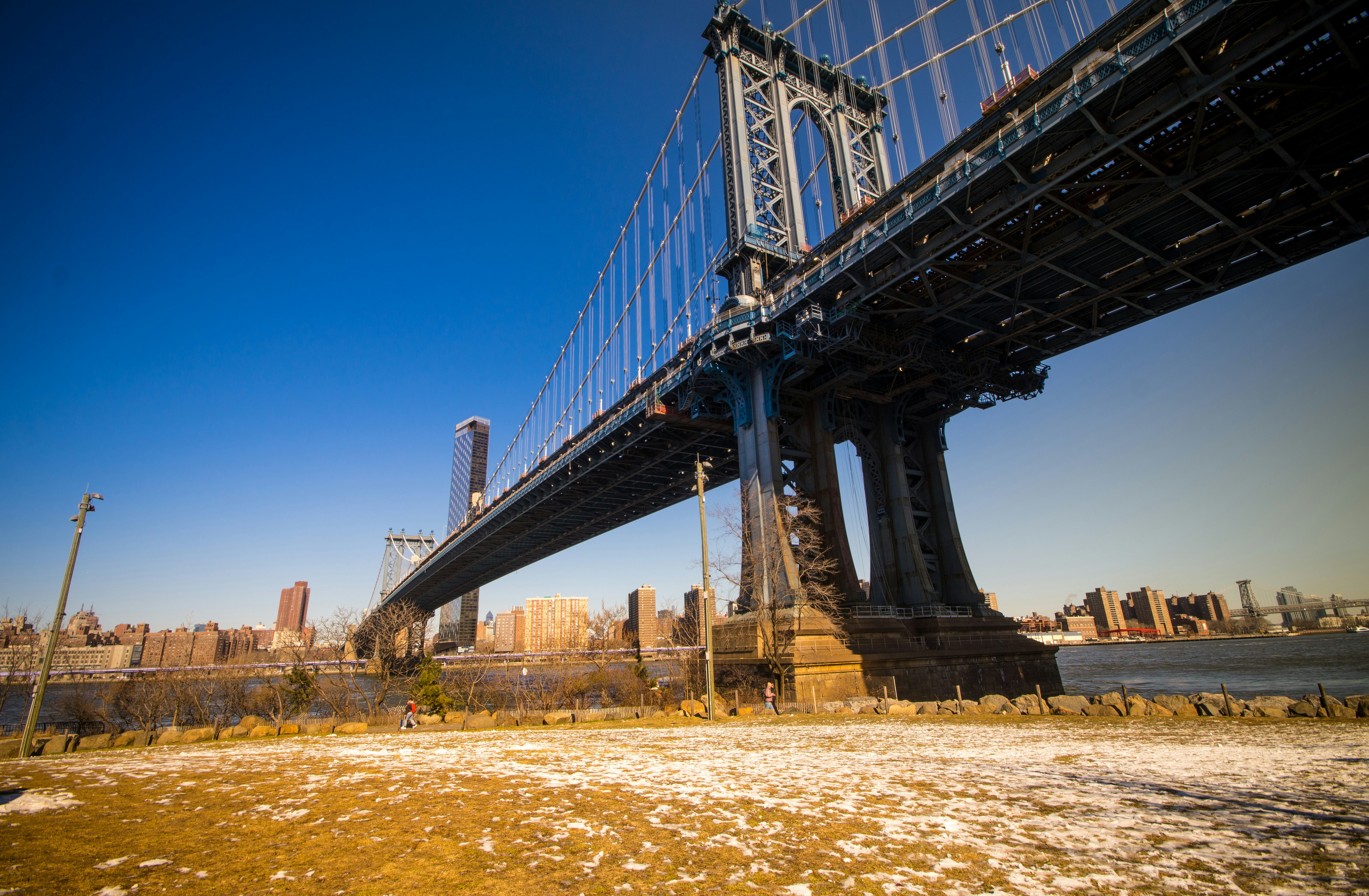Expansive steel bridge towering over a snowy riverside park with cityscape in the background.