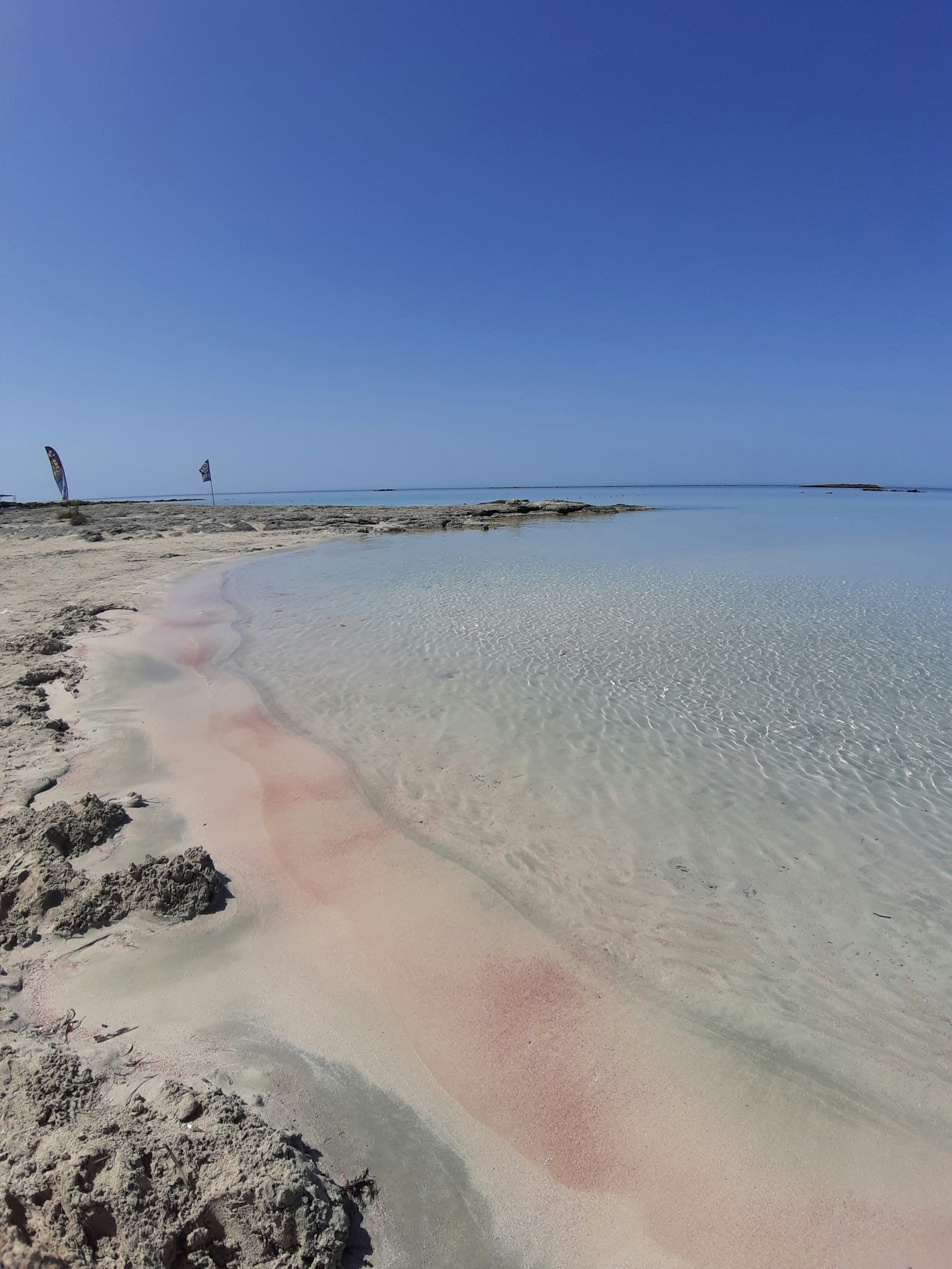 Gentle waves lap against a sandy beach, revealing a subtle pink hue along the shore under a clear blue sky.