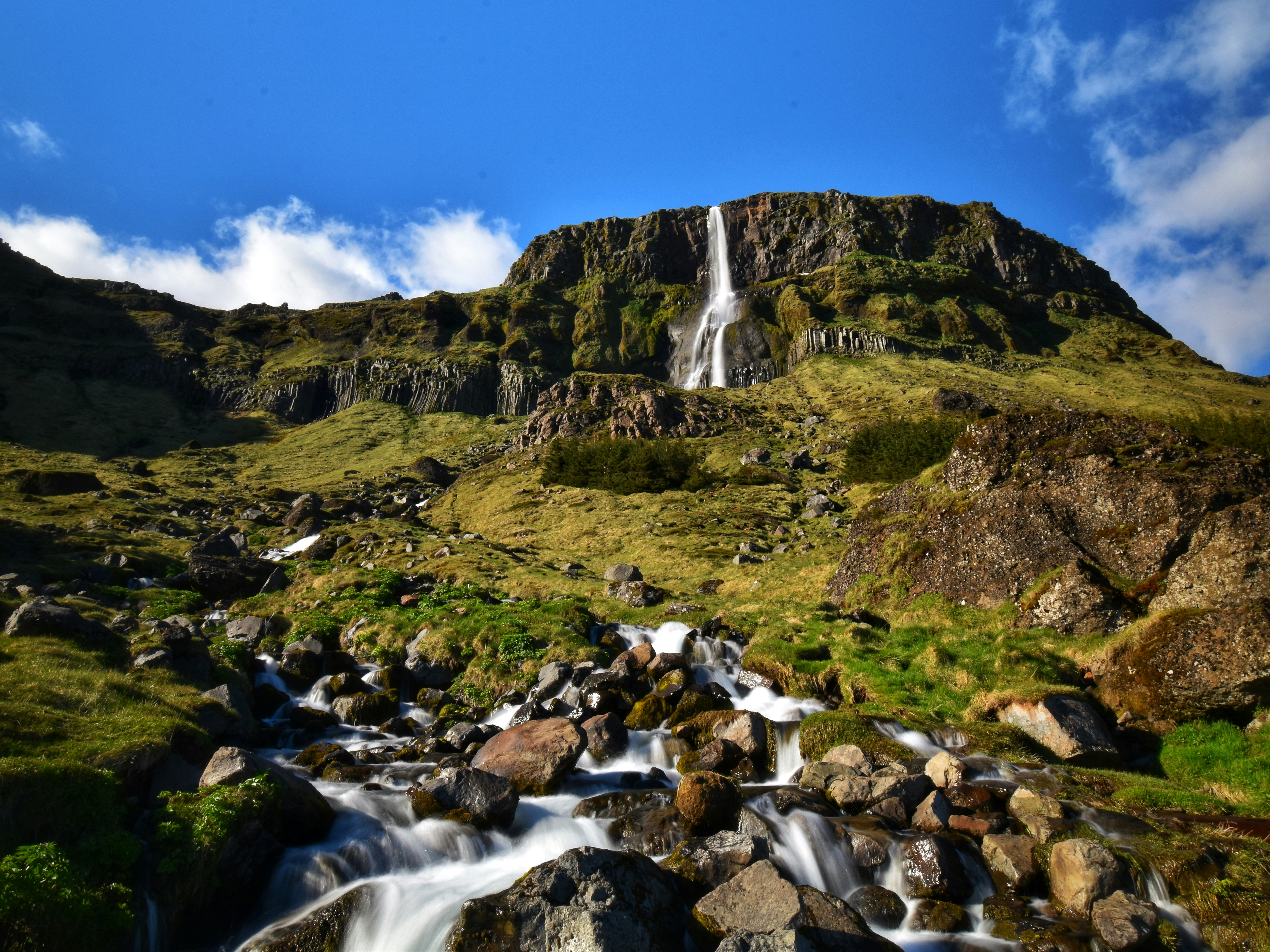 Waterfall flowing down a rocky, green hillside under a bright blue sky.