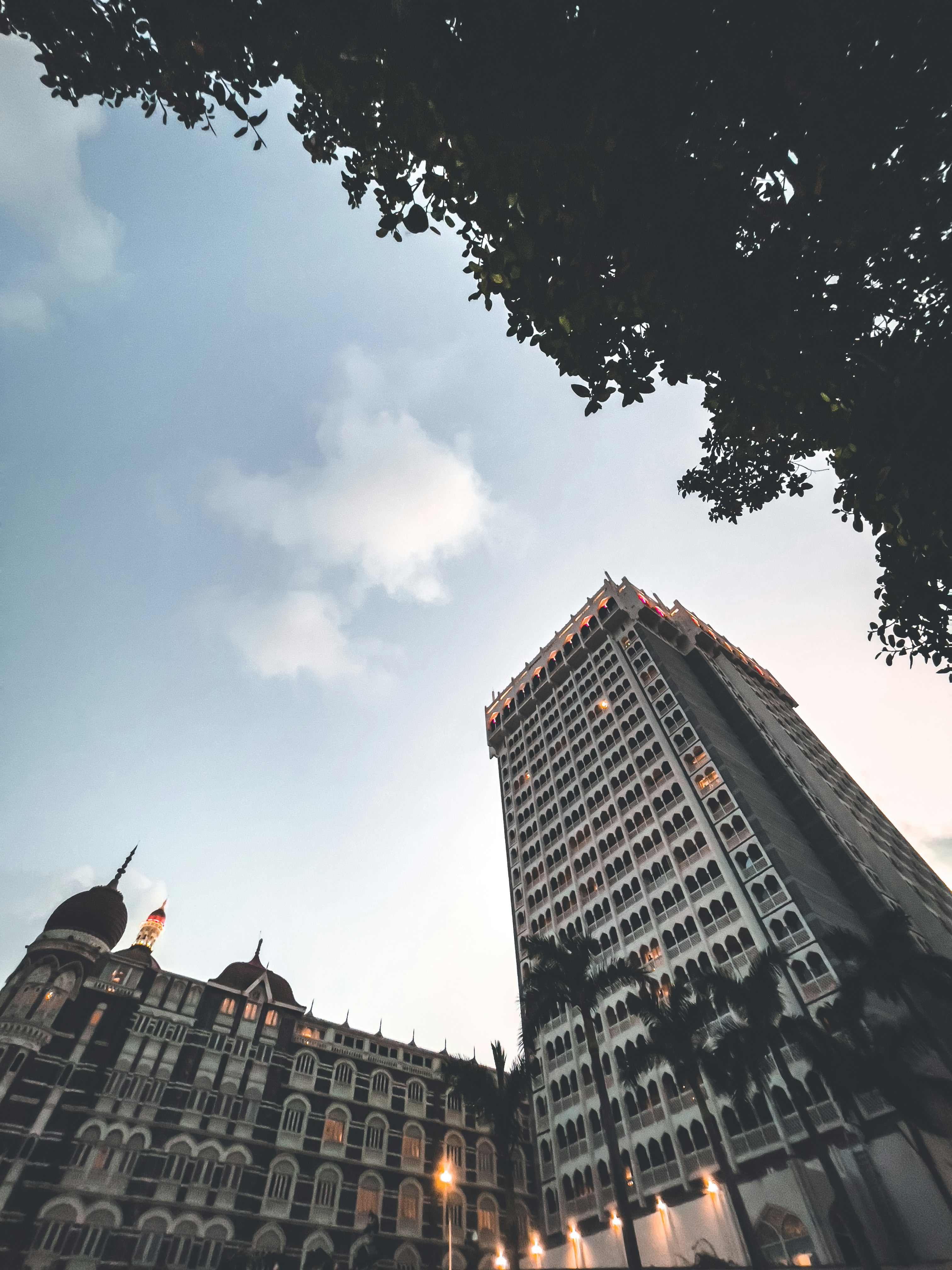 Majestic heritage hotel juxtaposed with a modern skyscraper under a twilight sky, framed by lush foliage.