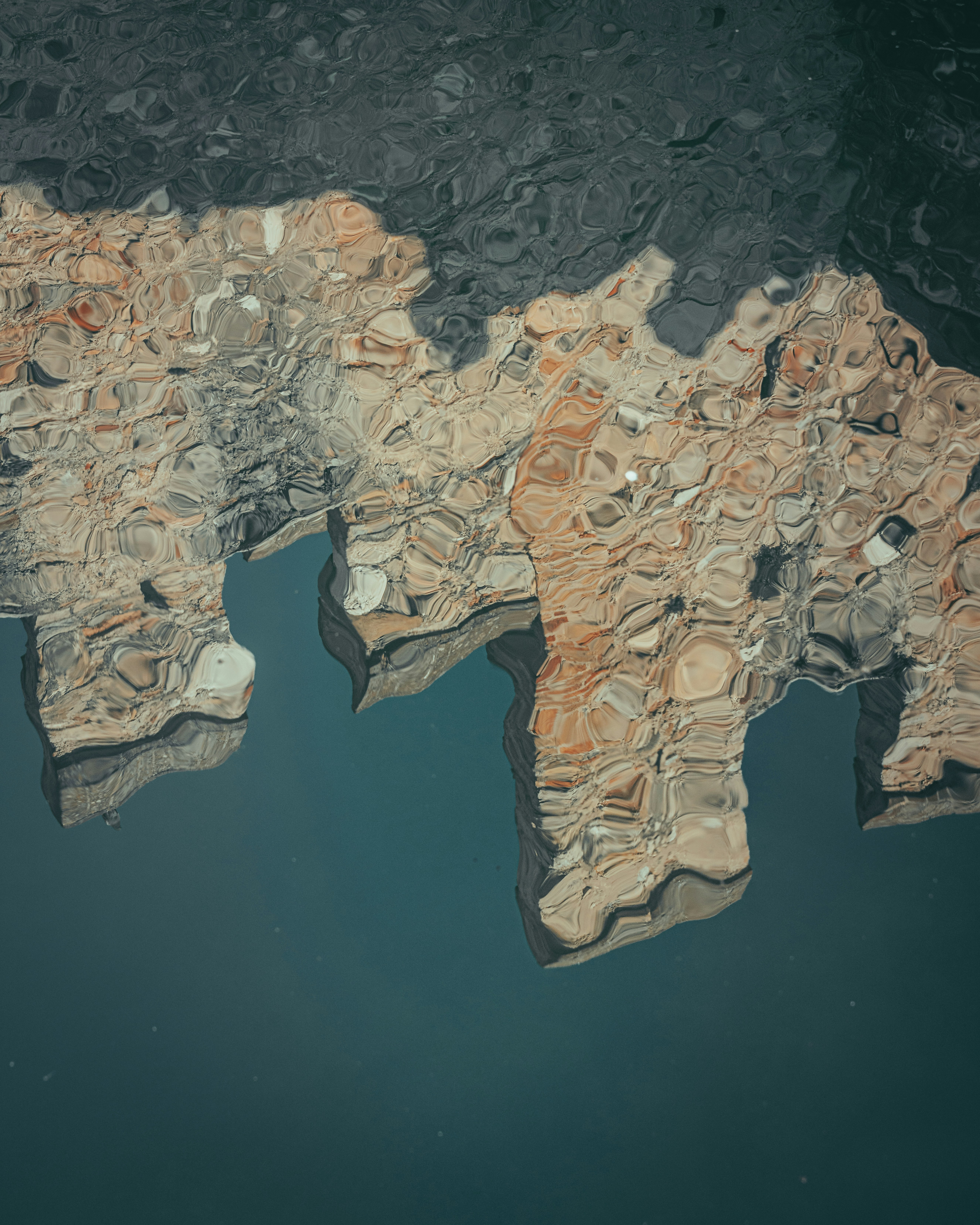 Rippling water reflects the textured patterns of a rocky shore, creating an abstract interplay of colors and shapes.