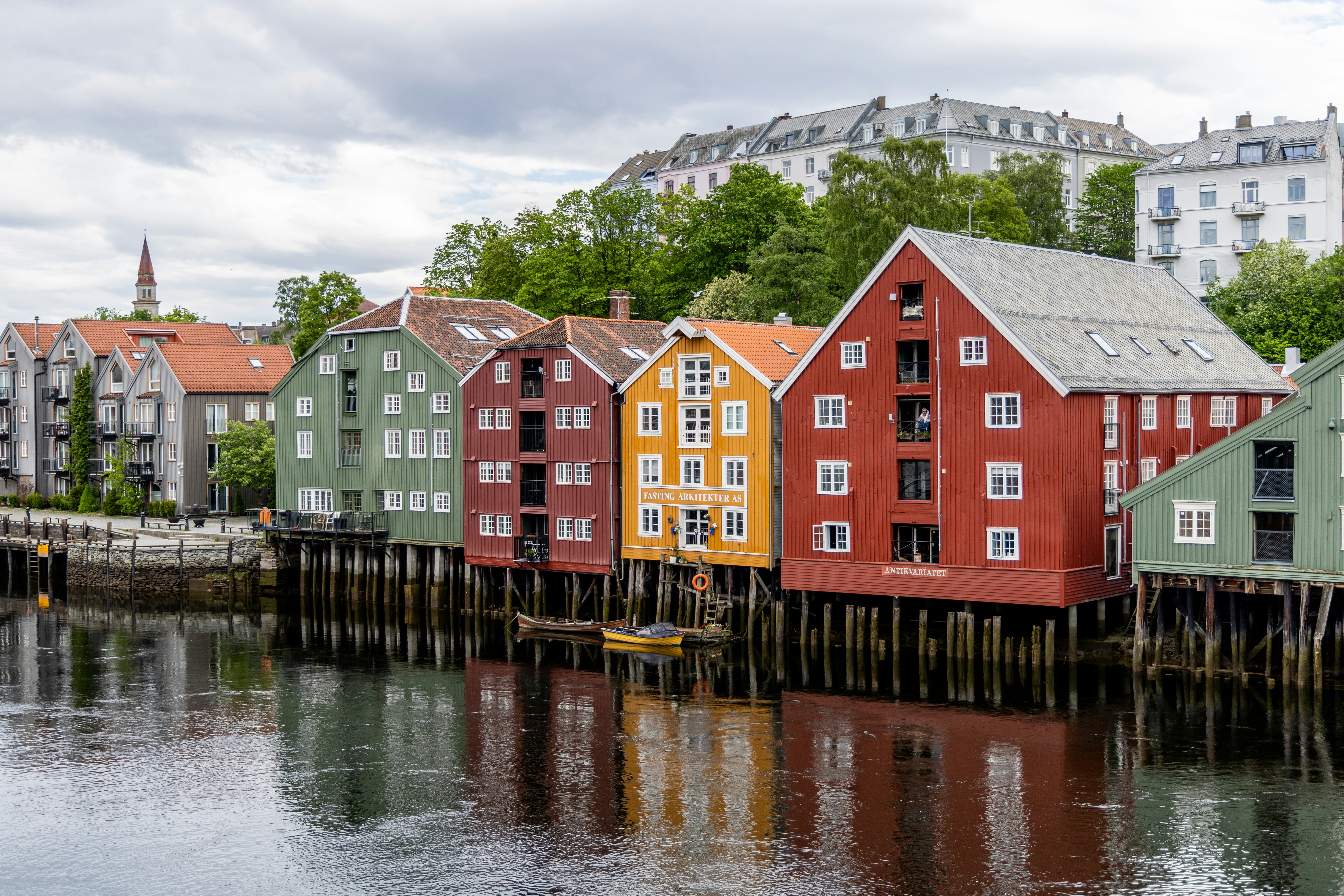 Colorful wooden buildings line the riverbank, reflected in the calm water under a cloudy sky.