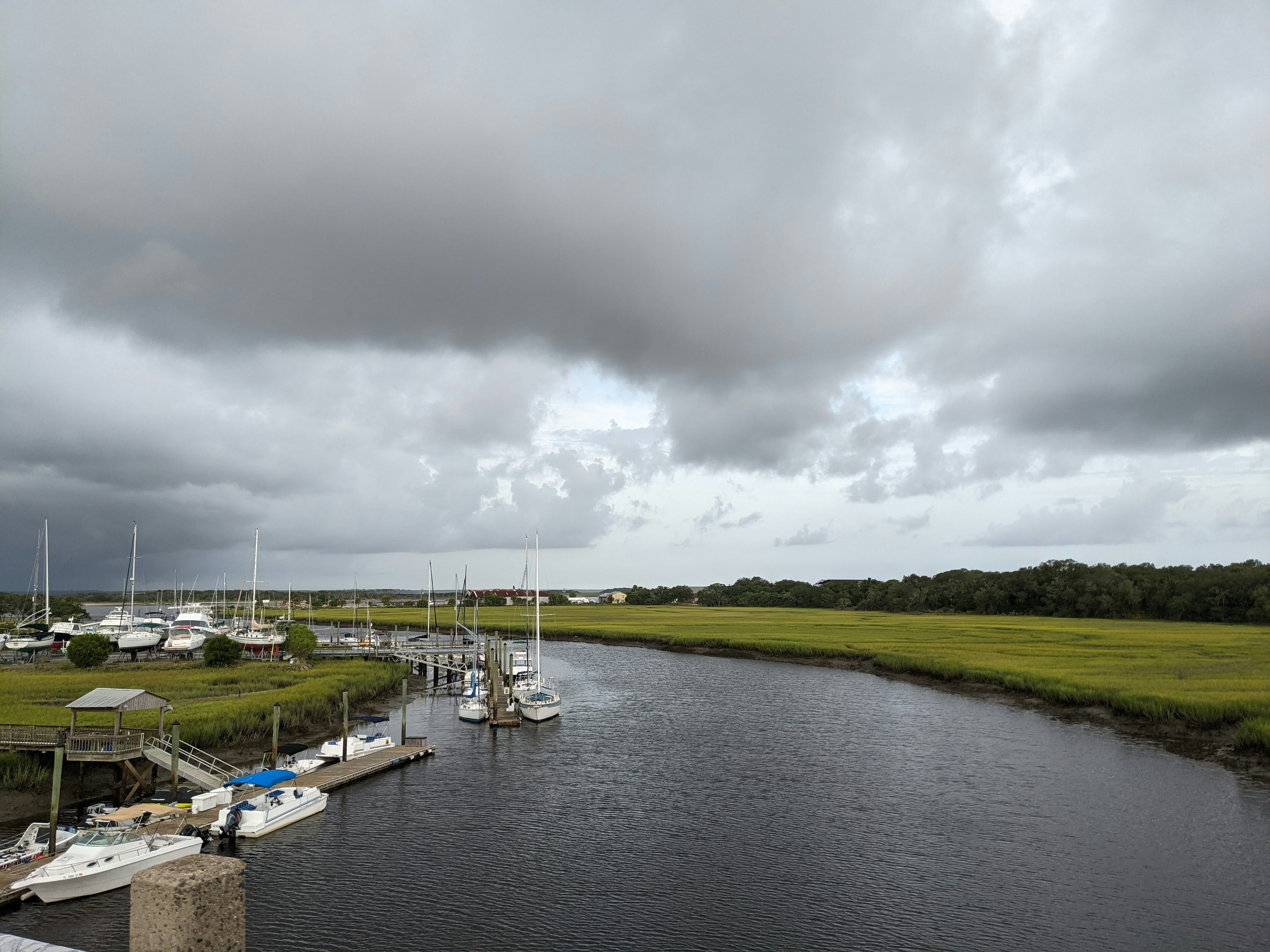 A tranquil marina scene with sailboats docked along a winding river, framed by lush green fields and dramatic clouds overhead.