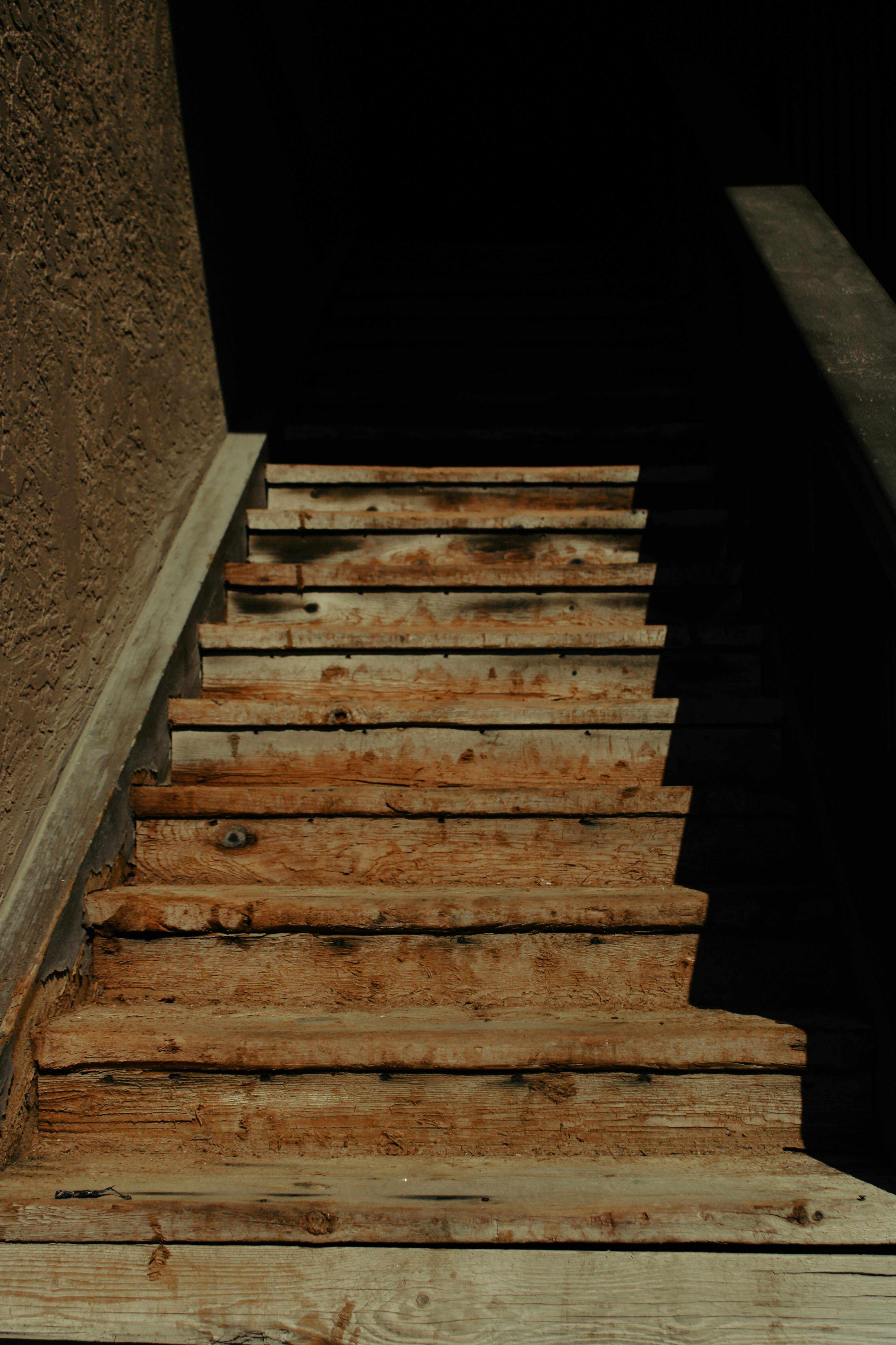 Worn wooden stairs lead down into a dimly lit space, showcasing the texture and age of the wood. Light plays on the surfaces, creating a stark contrast between light and shadow.