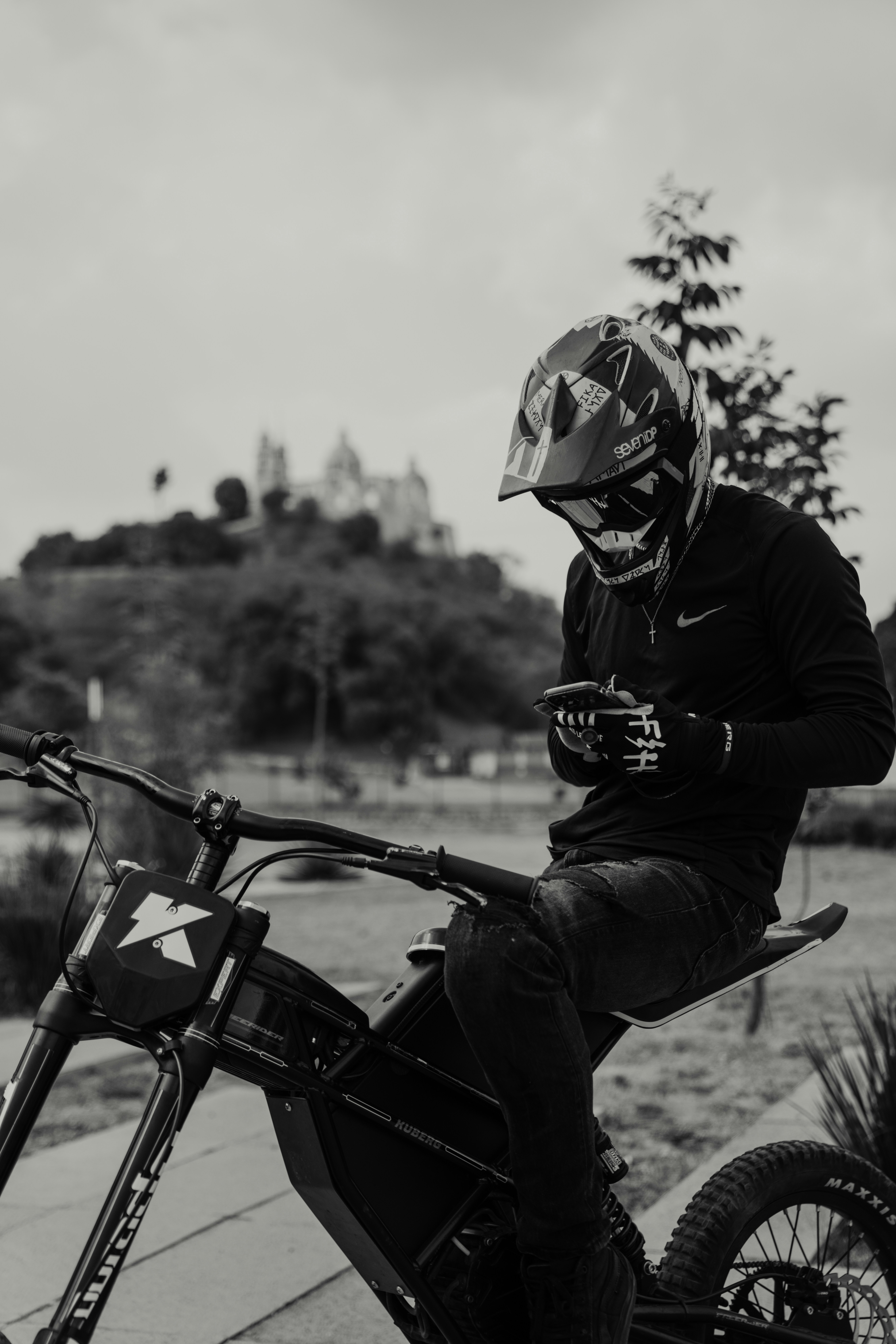 Person in a helmet sits on an electric bike, focused on a device, with a distant castle on a hill in the background.