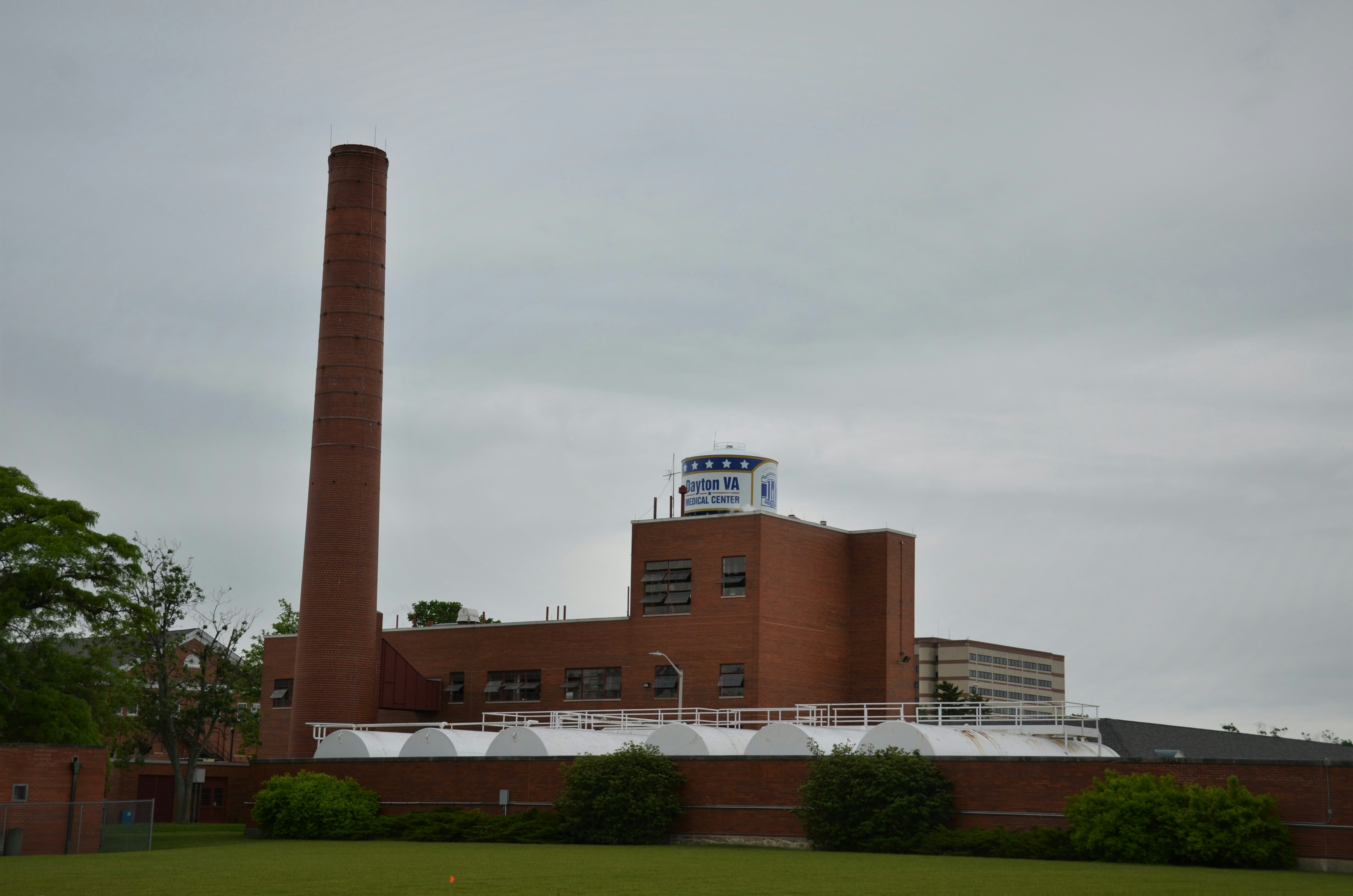 Brick industrial building with a tall smokestack and water tower, set against a cloudy sky. The structure reflects a bygone era of manufacturing.