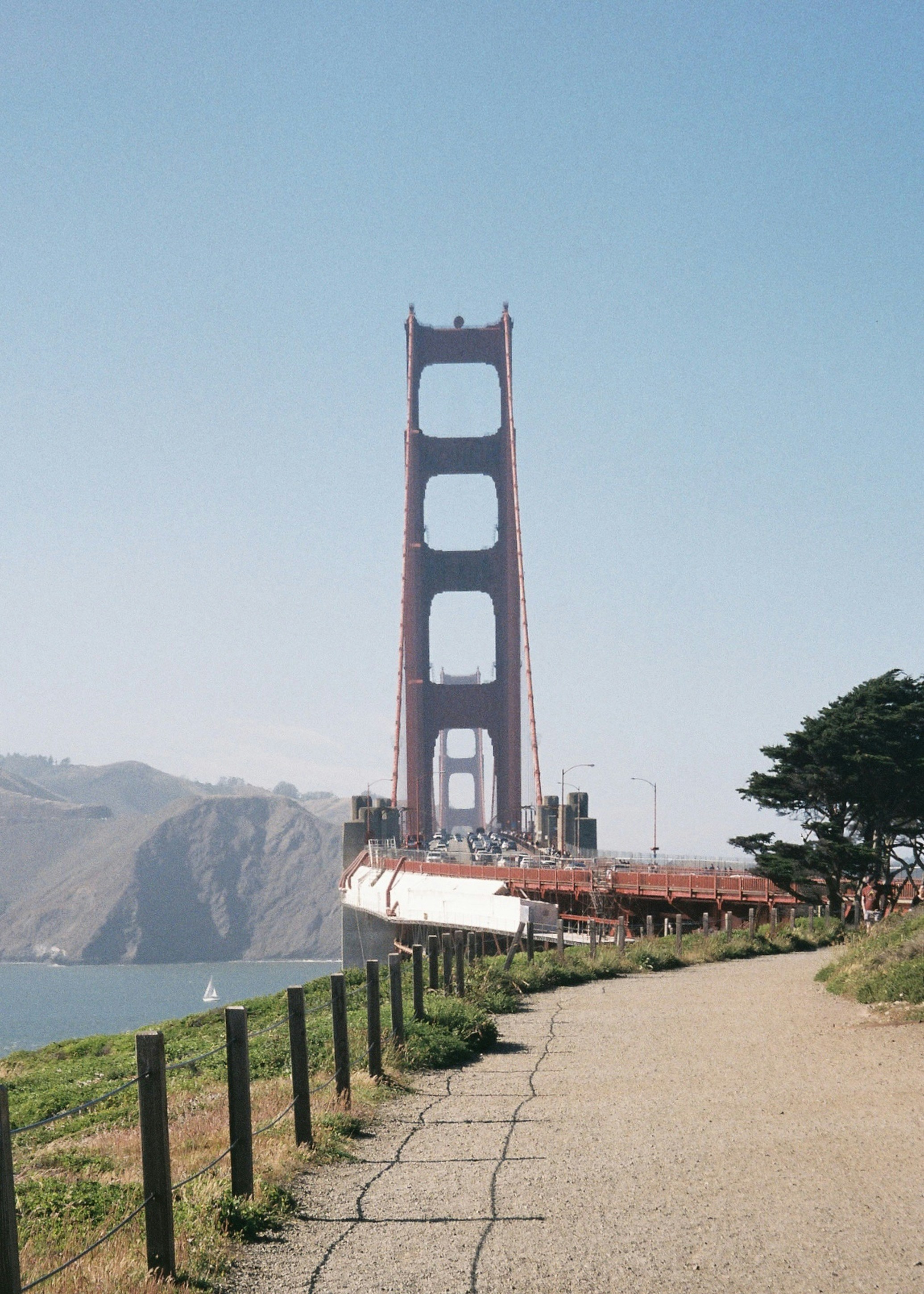 Golden Gate Bridge viewed from a scenic pathway, framed by lush greenery and distant hills. A sailboat glides across the bay, enhancing the tranquil atmosphere.