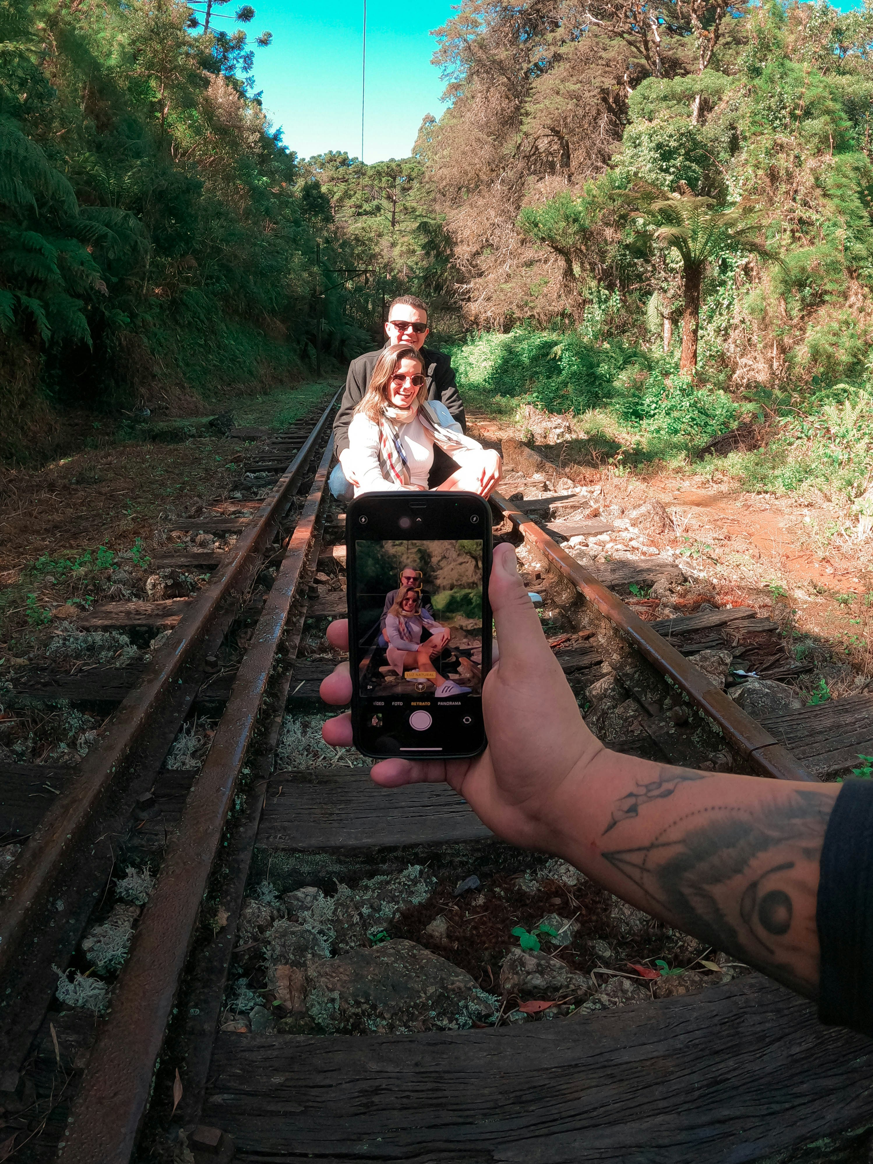 A hand holds a smartphone displaying a photo of a woman on overgrown railway tracks, with two people sitting behind her in a lush green setting.