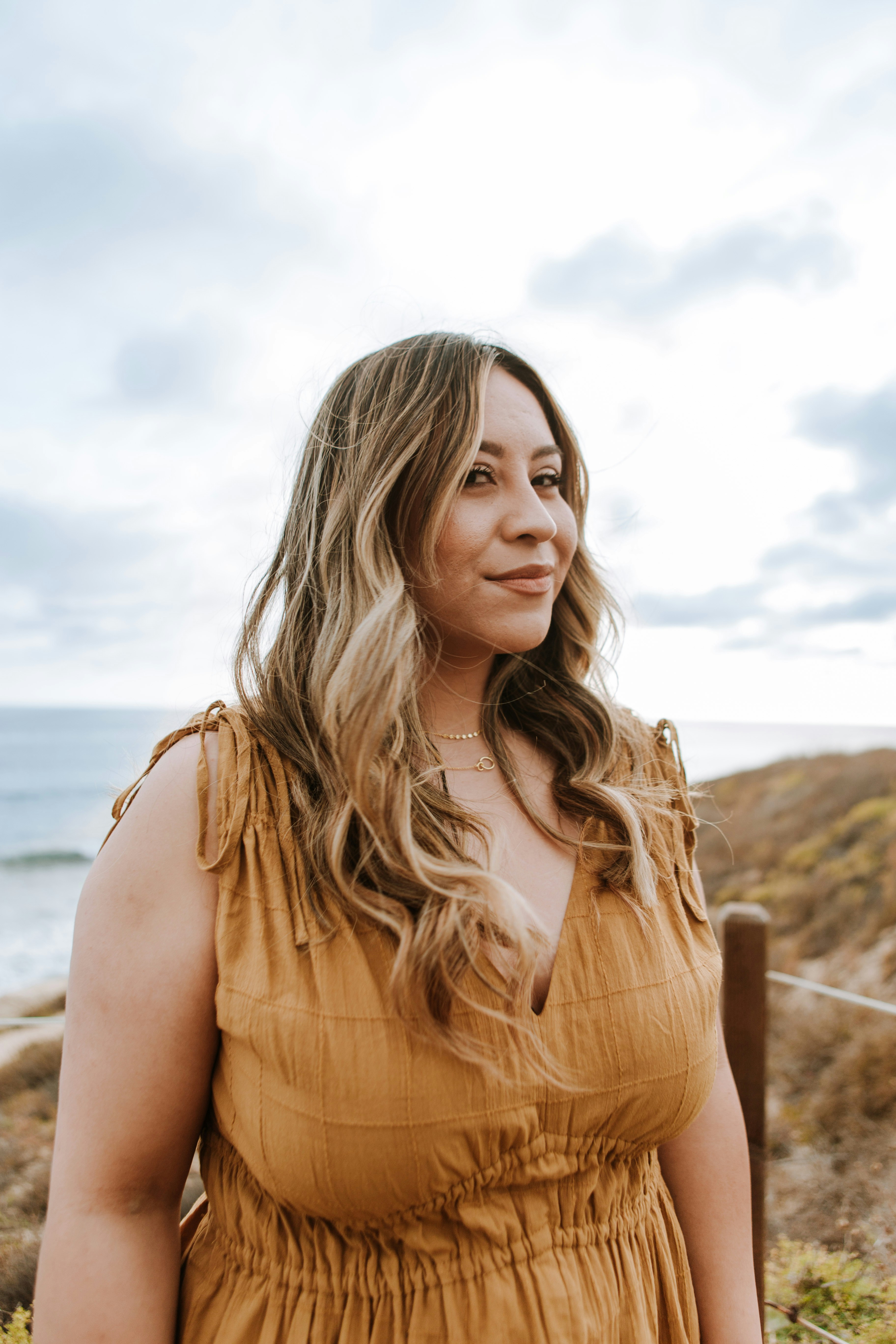 Woman in a golden dress poses with a serene expression against a coastal backdrop, capturing a moment of tranquility. 