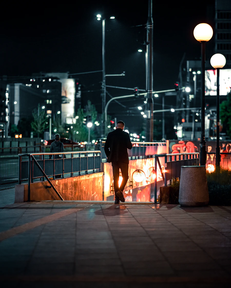 A man in a dark coat walks down steps into a bright underpass on a city street at night, with glowing lamps and traffic lights in the background.