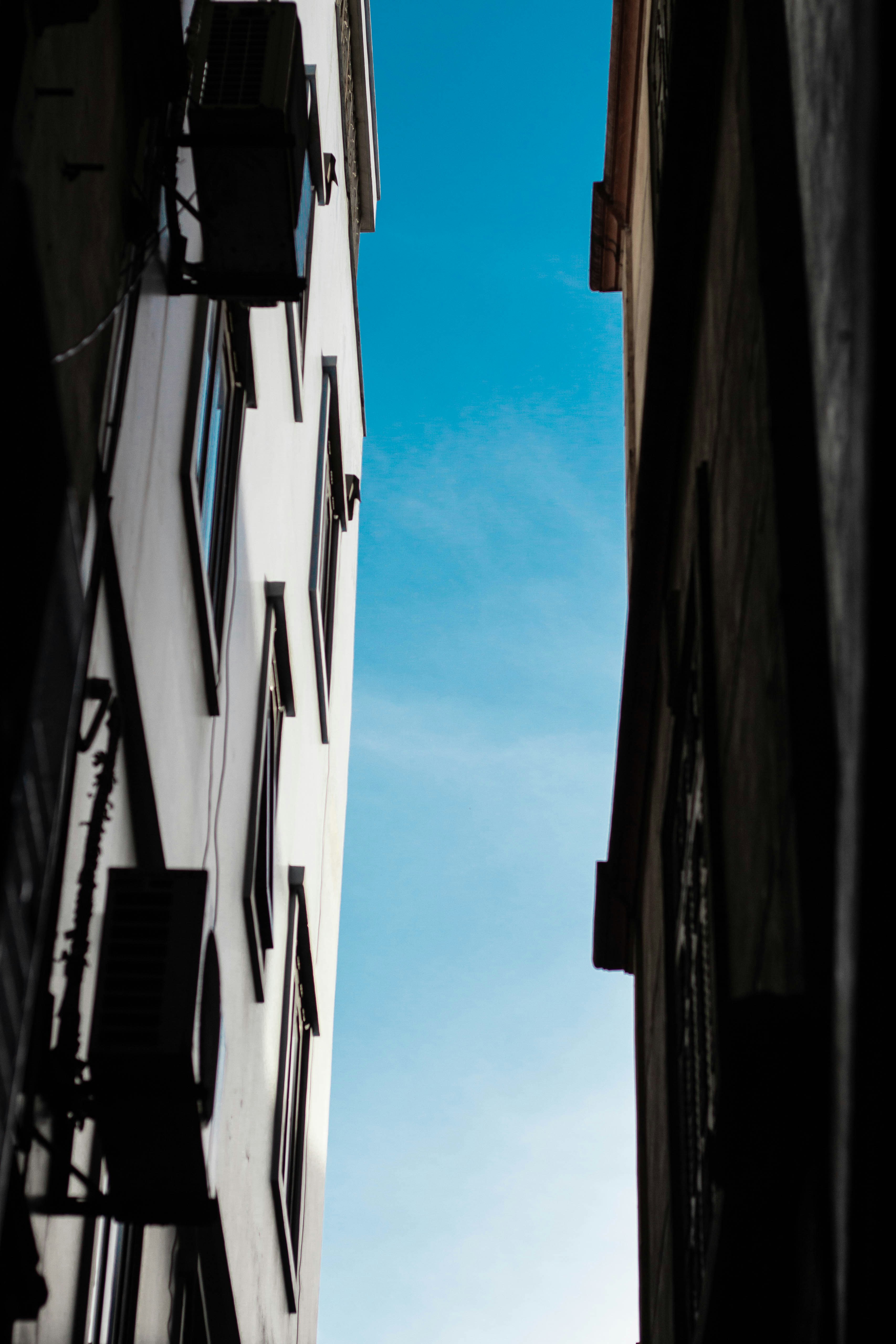 Narrow alleyway framed by tall buildings, revealing a patch of blue sky above. Air conditioning units hang from the walls, adding to the urban atmosphere.