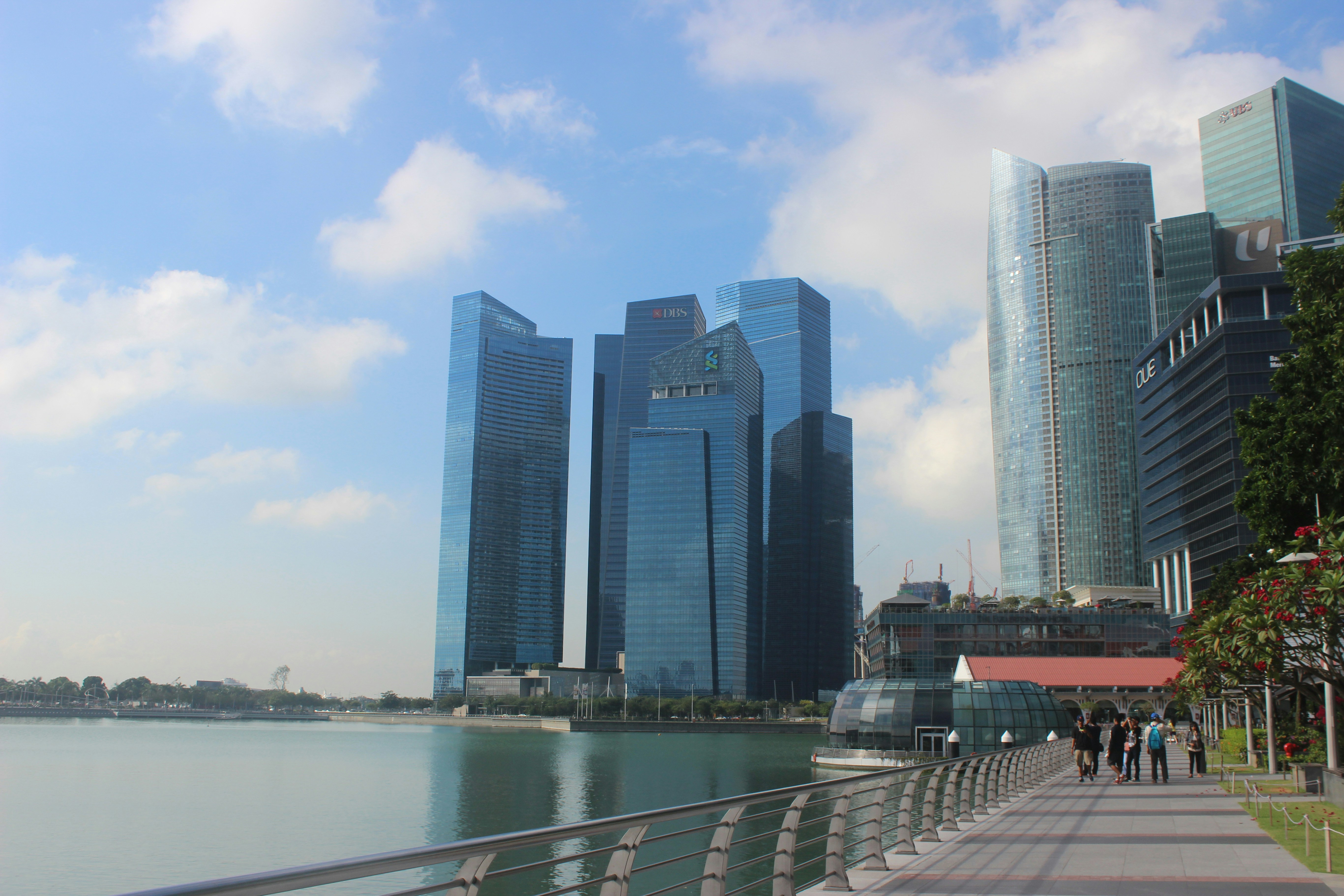 Modern skyscrapers reflecting in calm waters, framed by a scenic promenade and clear skies.