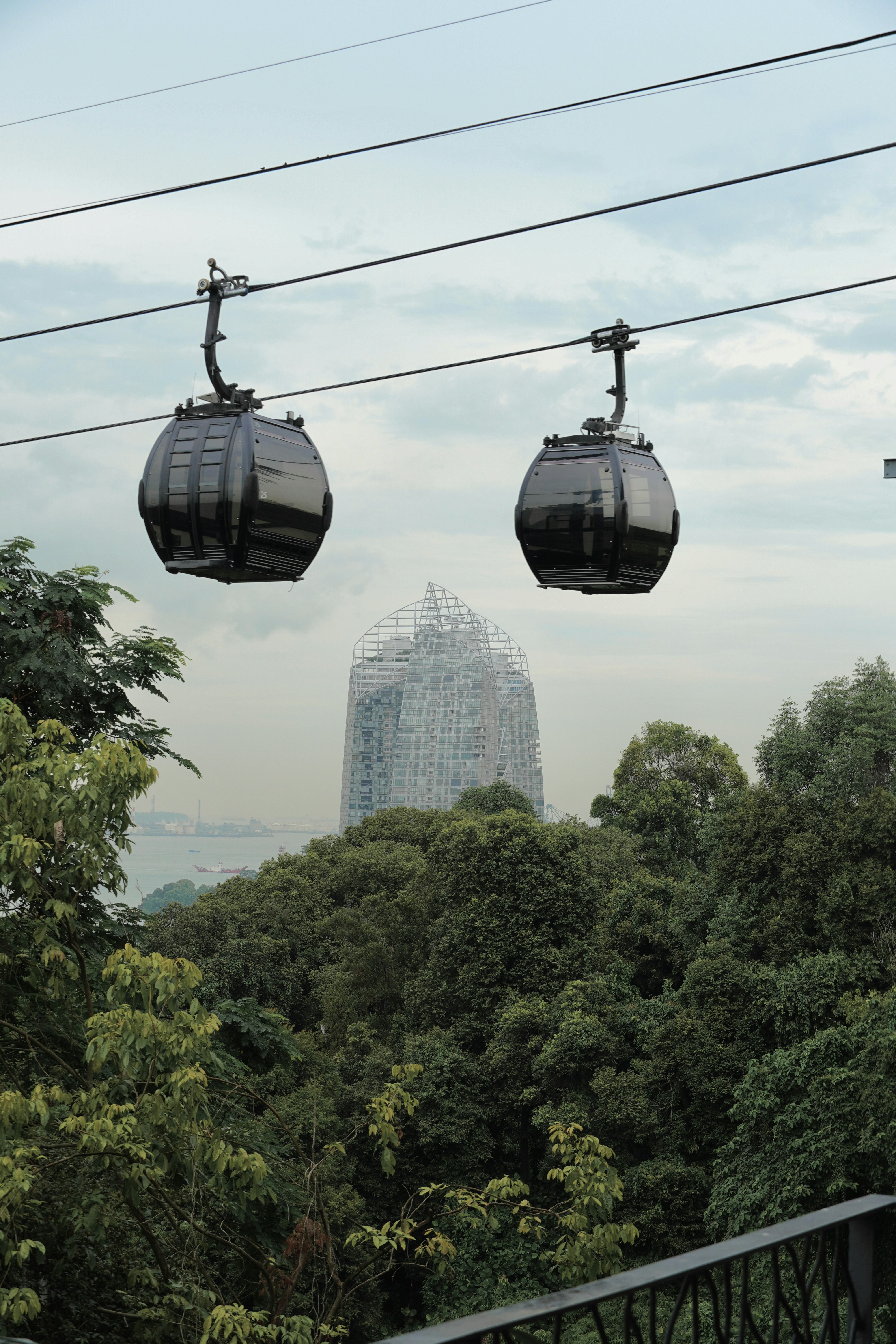 Two black cable cars glide above lush greenery with a partially constructed modern building in the background. The scene captures the blend of nature and urban development.