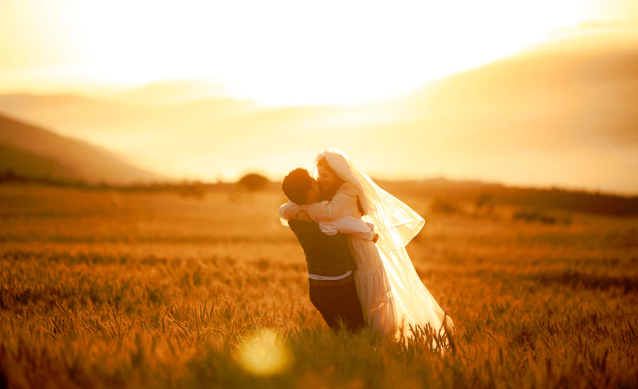 A bride and groom embrace in a sunlit field at golden hour, with warm backlight and a glowing veil. Rolling hills fade softly into the bright horizon.