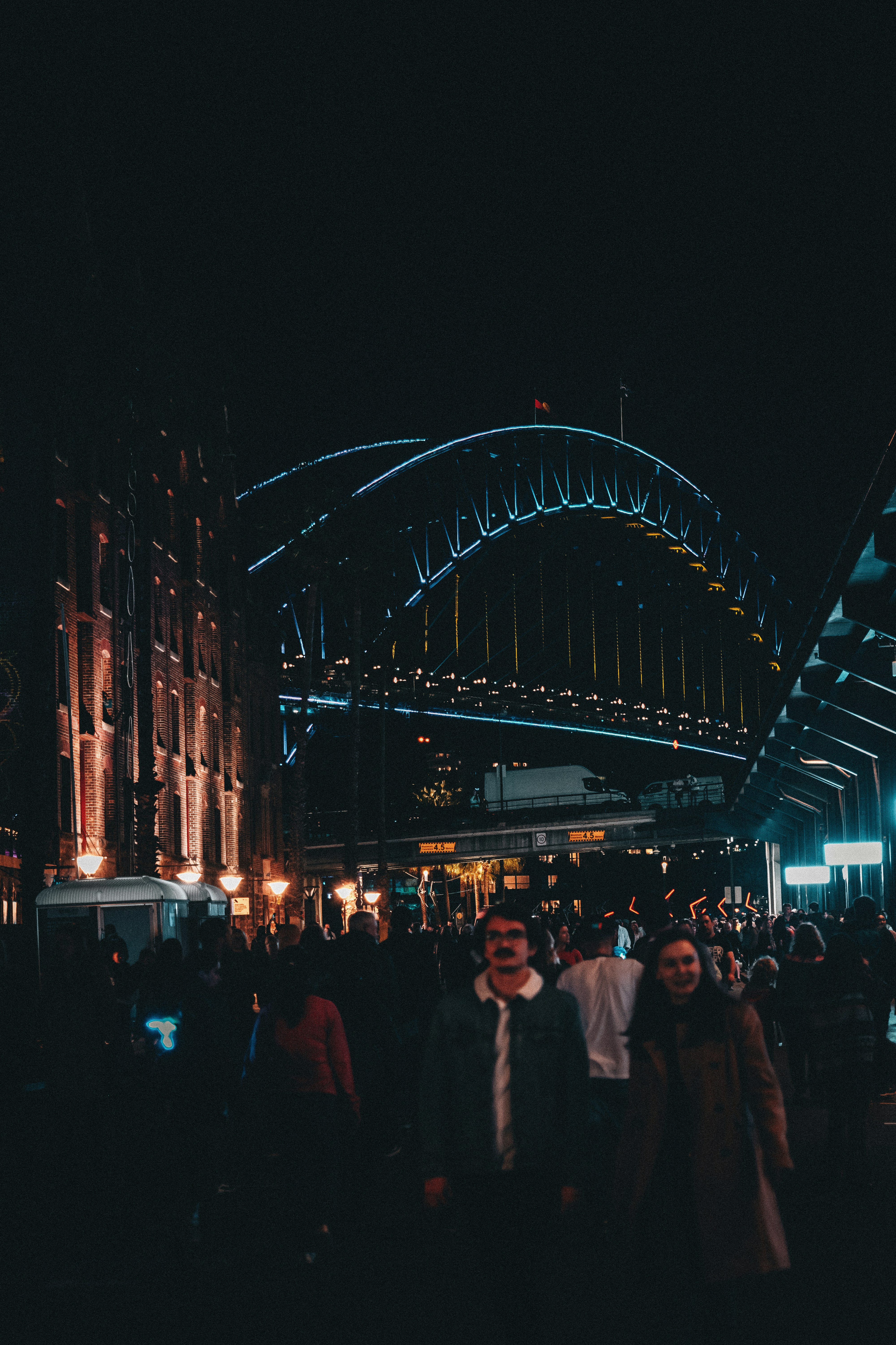 Crowd gathered at night, illuminated by colorful lights from the bridge above. The scene captures the lively atmosphere of an urban gathering.