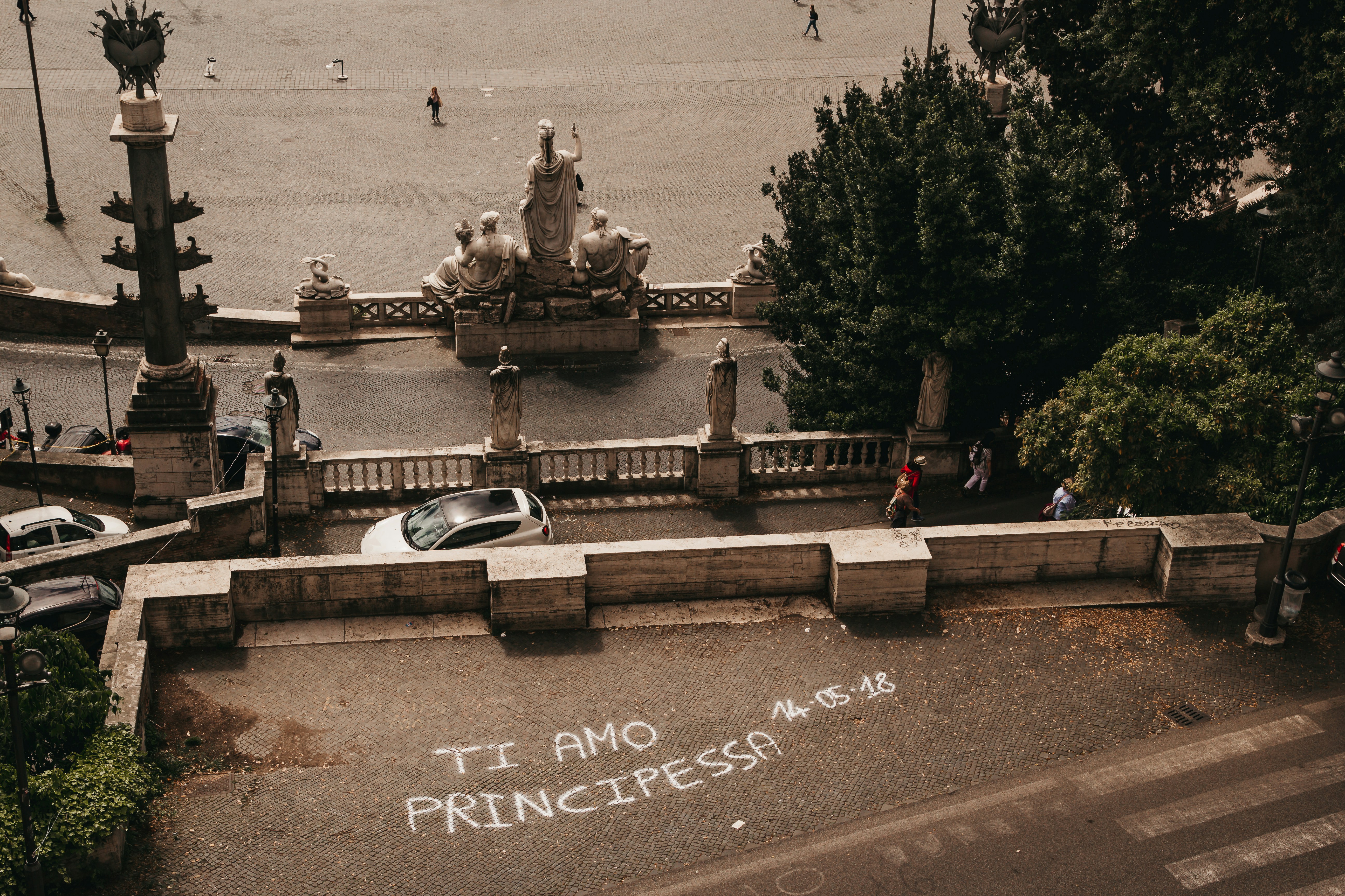Graffiti reading 'TI AMO PRINCIPESSA' on a stone wall, surrounded by urban scenery and greenery. A moment of romance captured in a bustling environment.