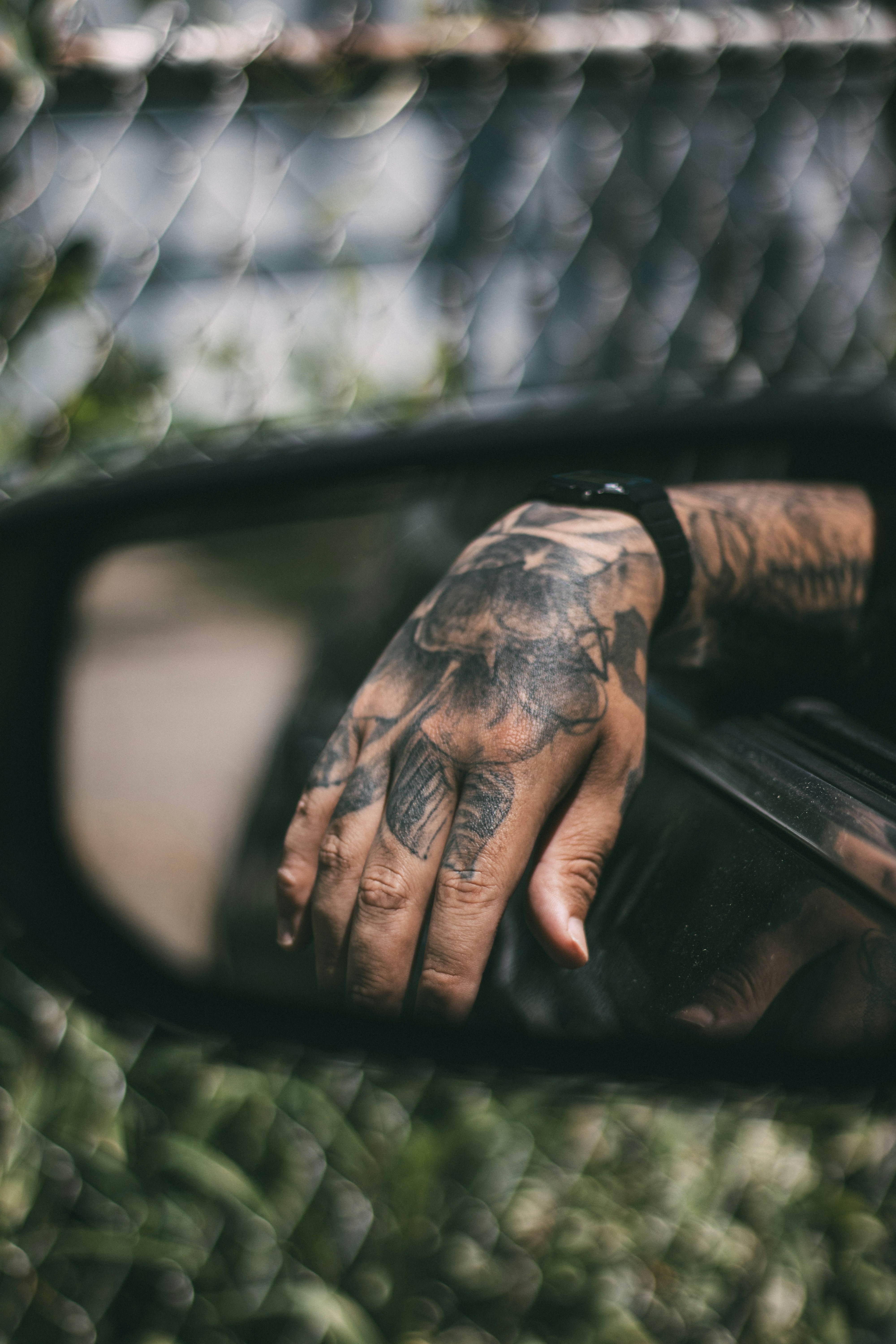 Tattooed hand resting on a car door, captured through a side mirror with a blurred background of greenery and chain-link fence.