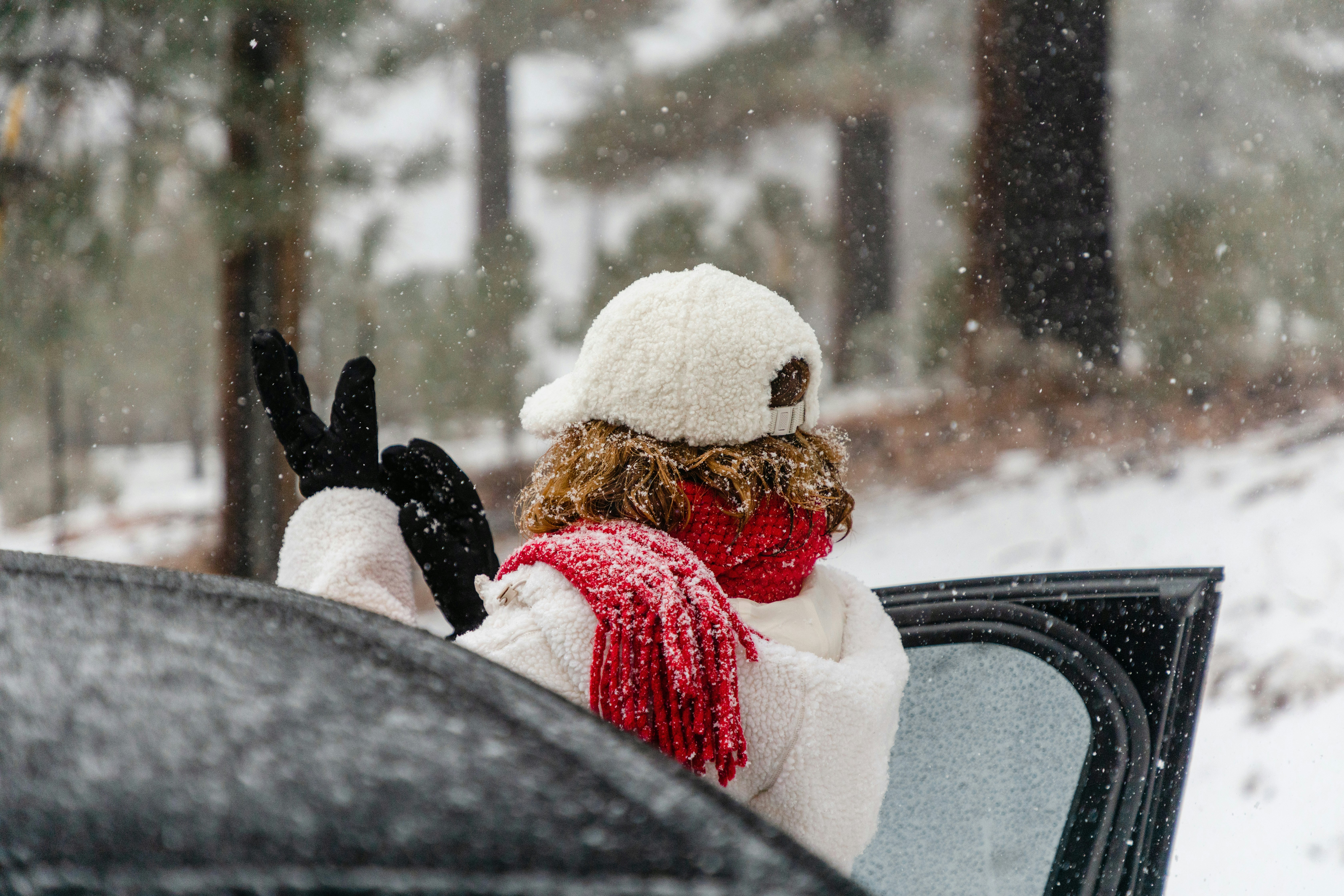 A person with curly hair wearing a white hat and red scarf playfully gestures peace while snow falls around them, creating a festive winter atmosphere.