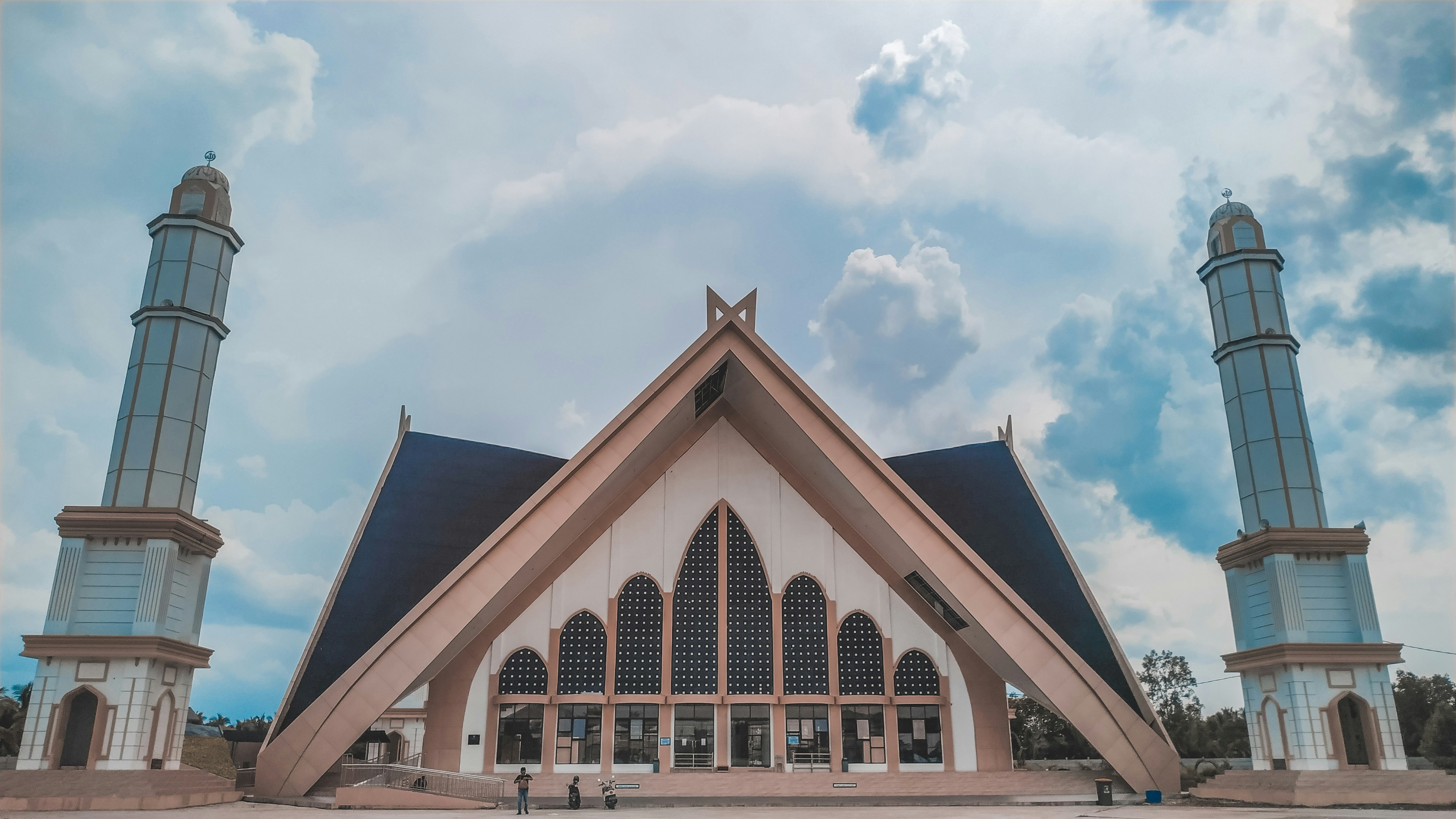 Modern mosque with triangular roof and two tall minarets under a partly cloudy sky.