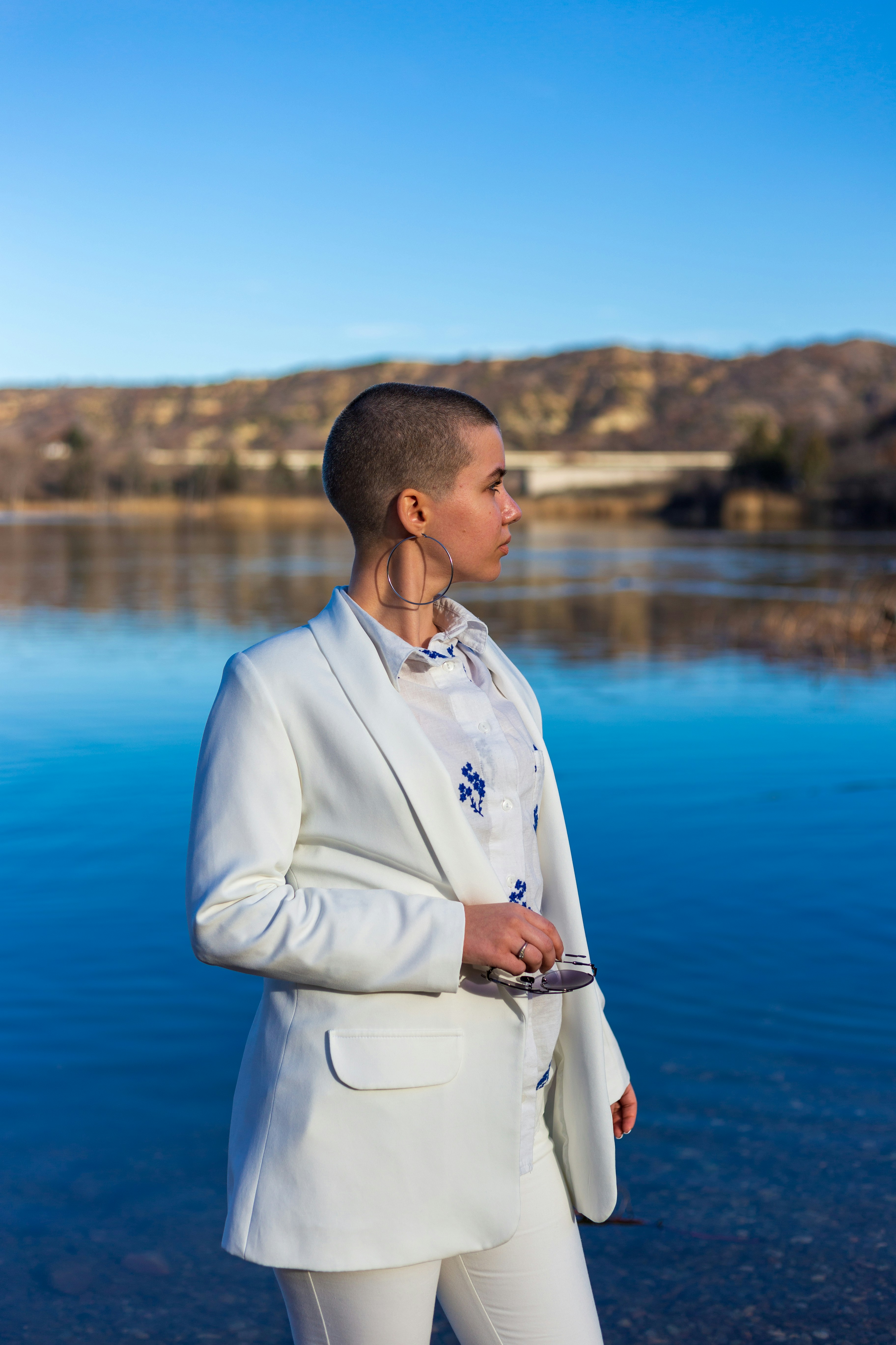 A person in a white suit stands by a tranquil lake, gazing thoughtfully into the distance. The serene water reflects the surrounding landscape.