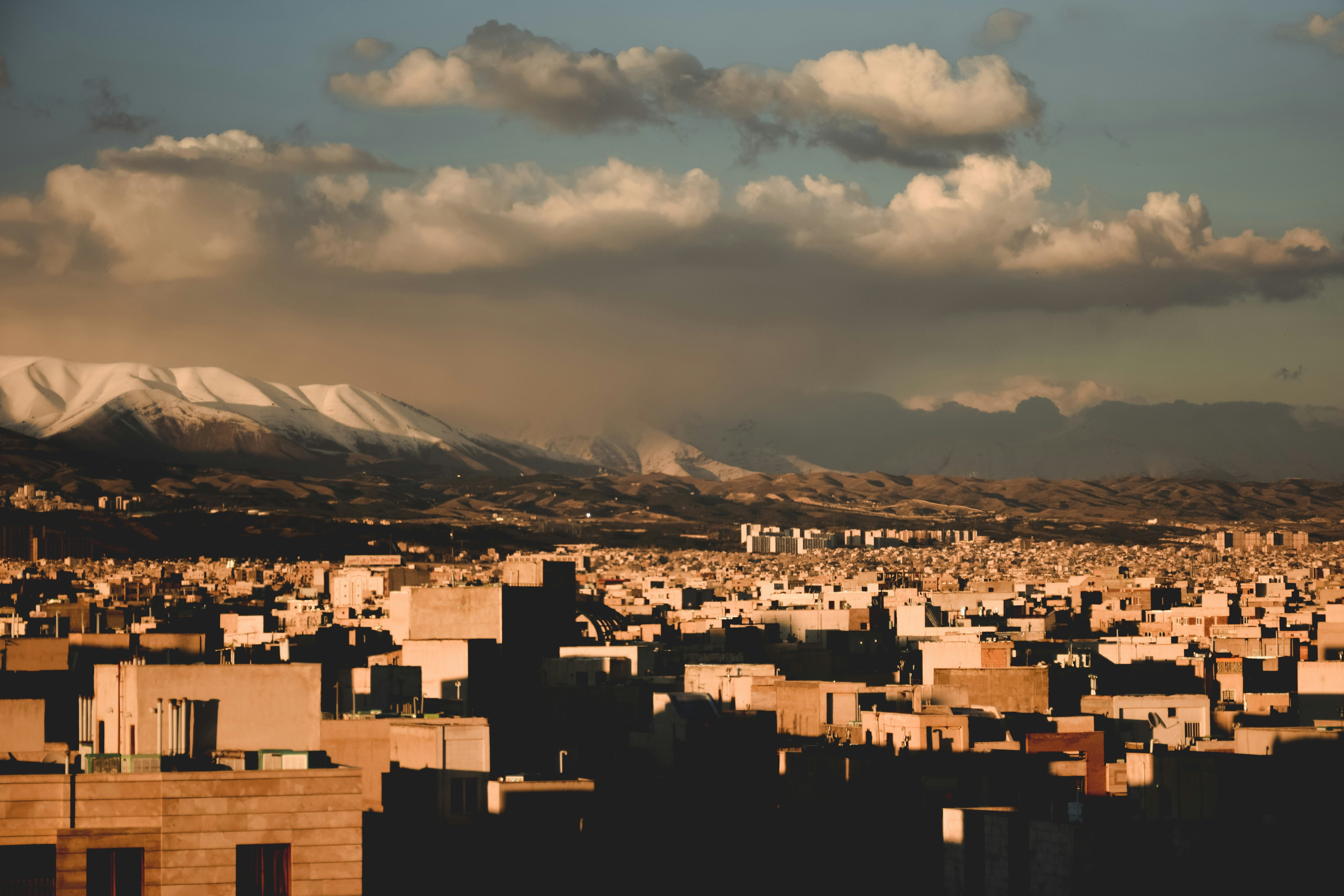 A sprawling cityscape with layered buildings in the foreground, set against dramatic snow-capped mountains and a dynamic sky.