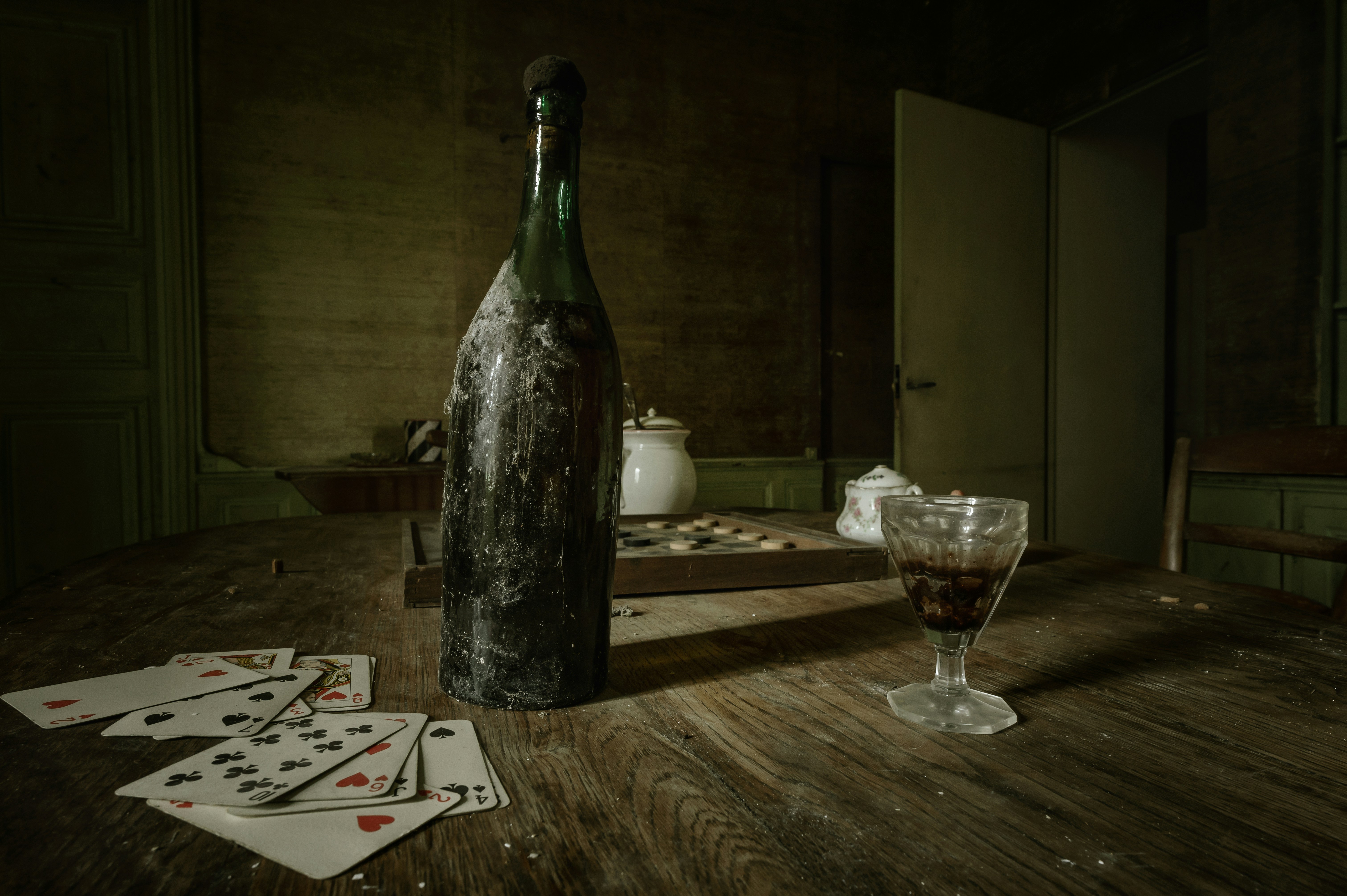 Dusty bottle and glass sit on a weathered table surrounded by scattered playing cards, evoking memories of past gatherings. 
