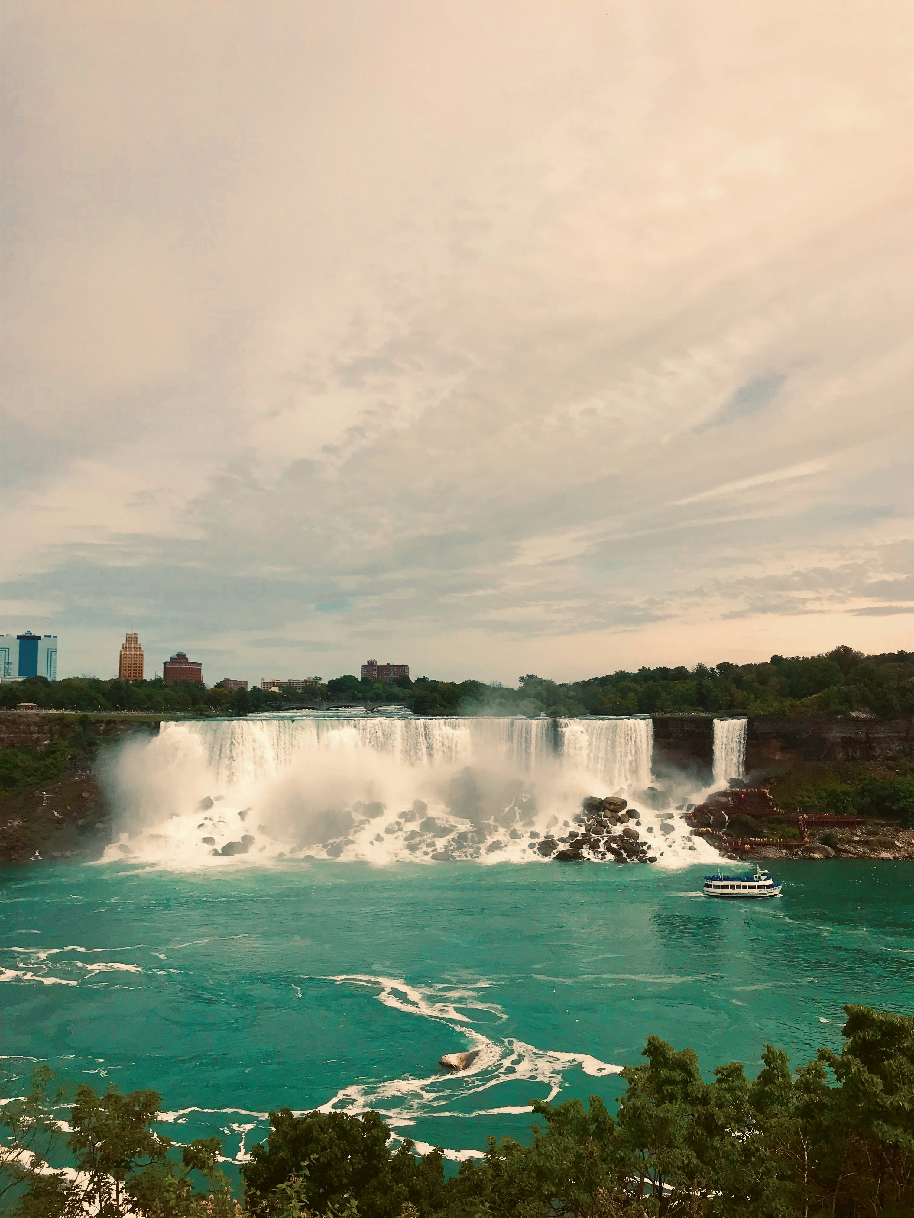 Powerful waterfalls plunging into turquoise waters, framed by lush greenery and distant city skyline.