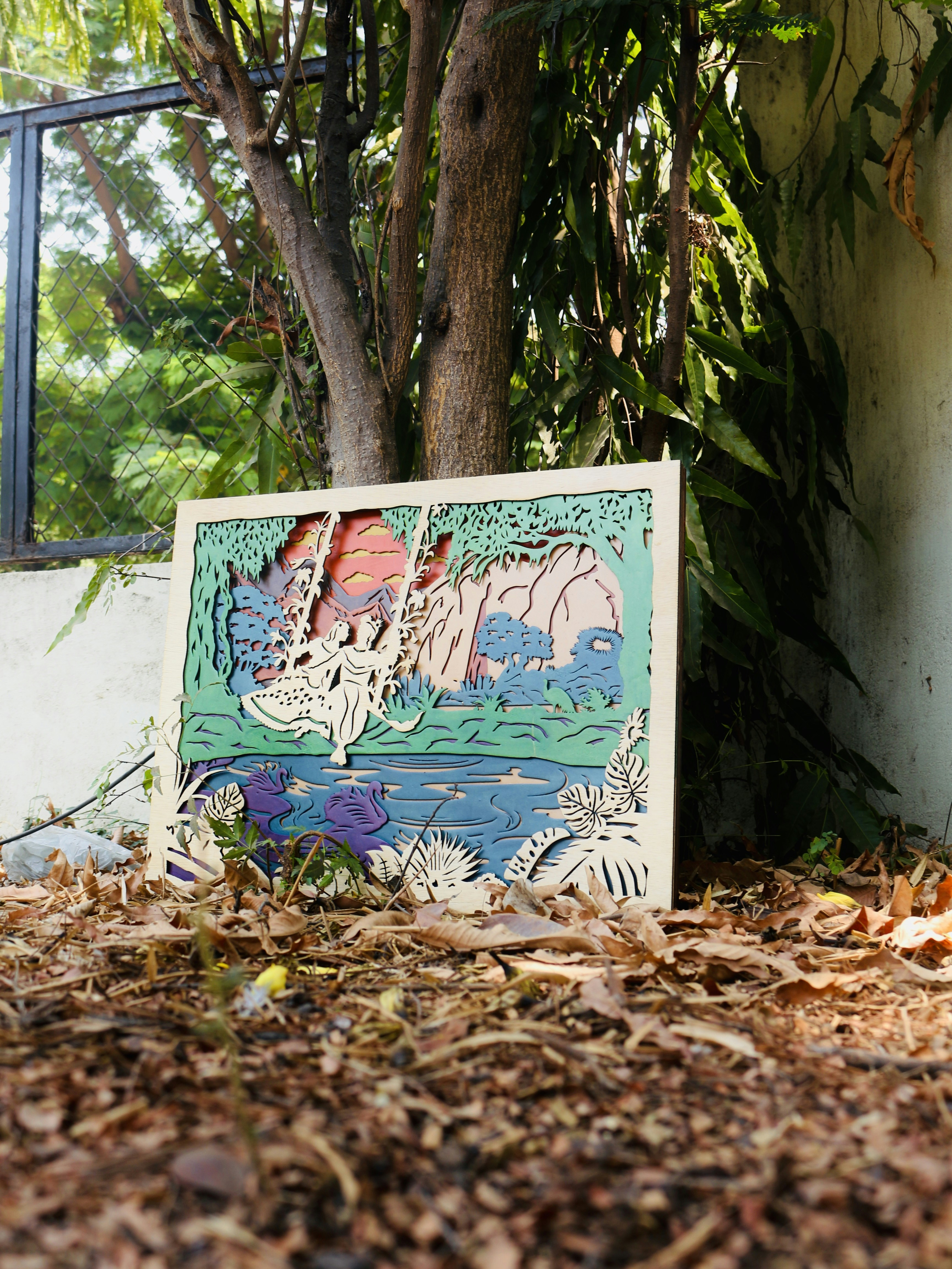Papercut Forest Scene: dye-cut art resting among leaves beneath a shadowy garden wall
