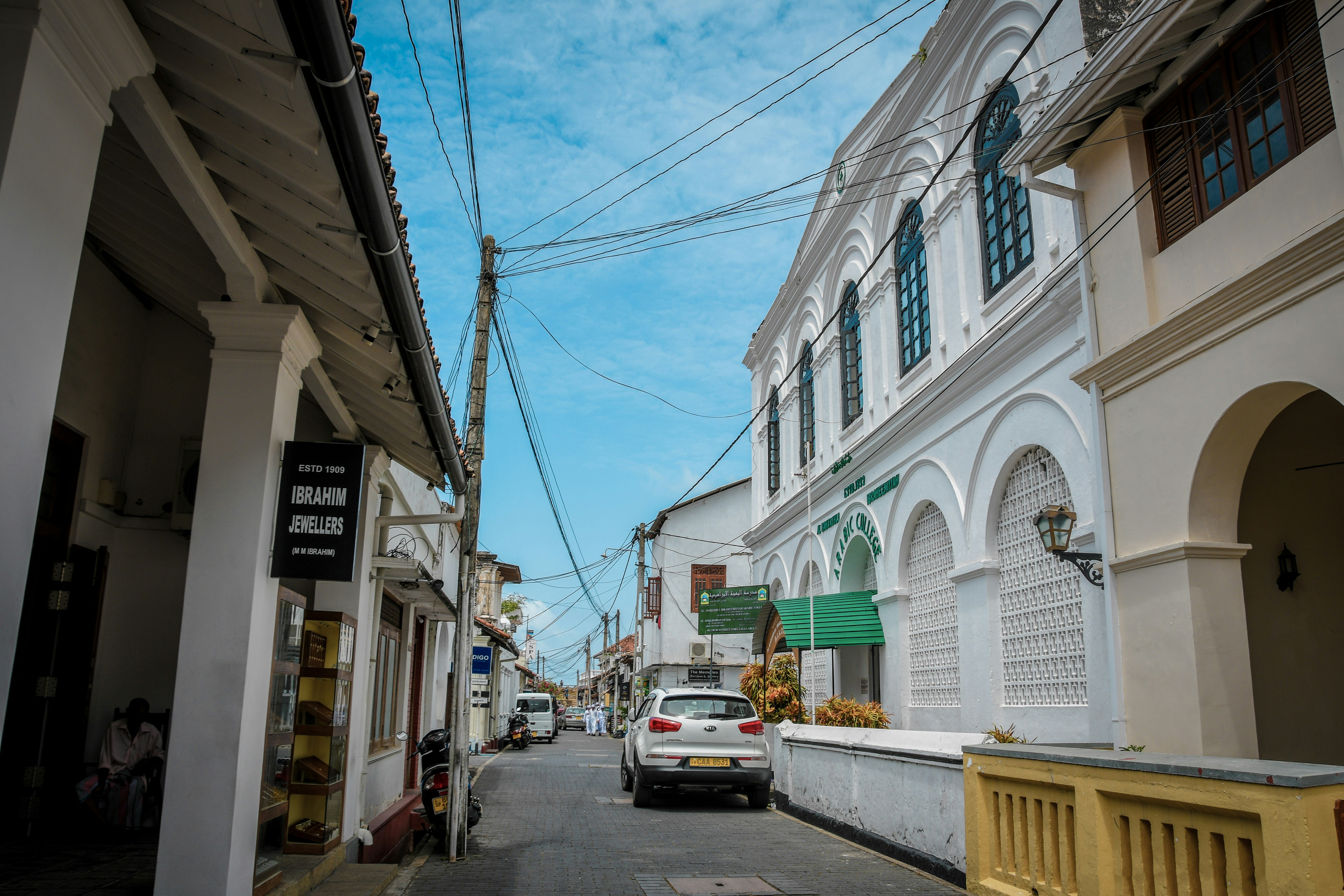 Car parked alongside a narrow street flanked by white colonial-style buildings under a clear blue sky.