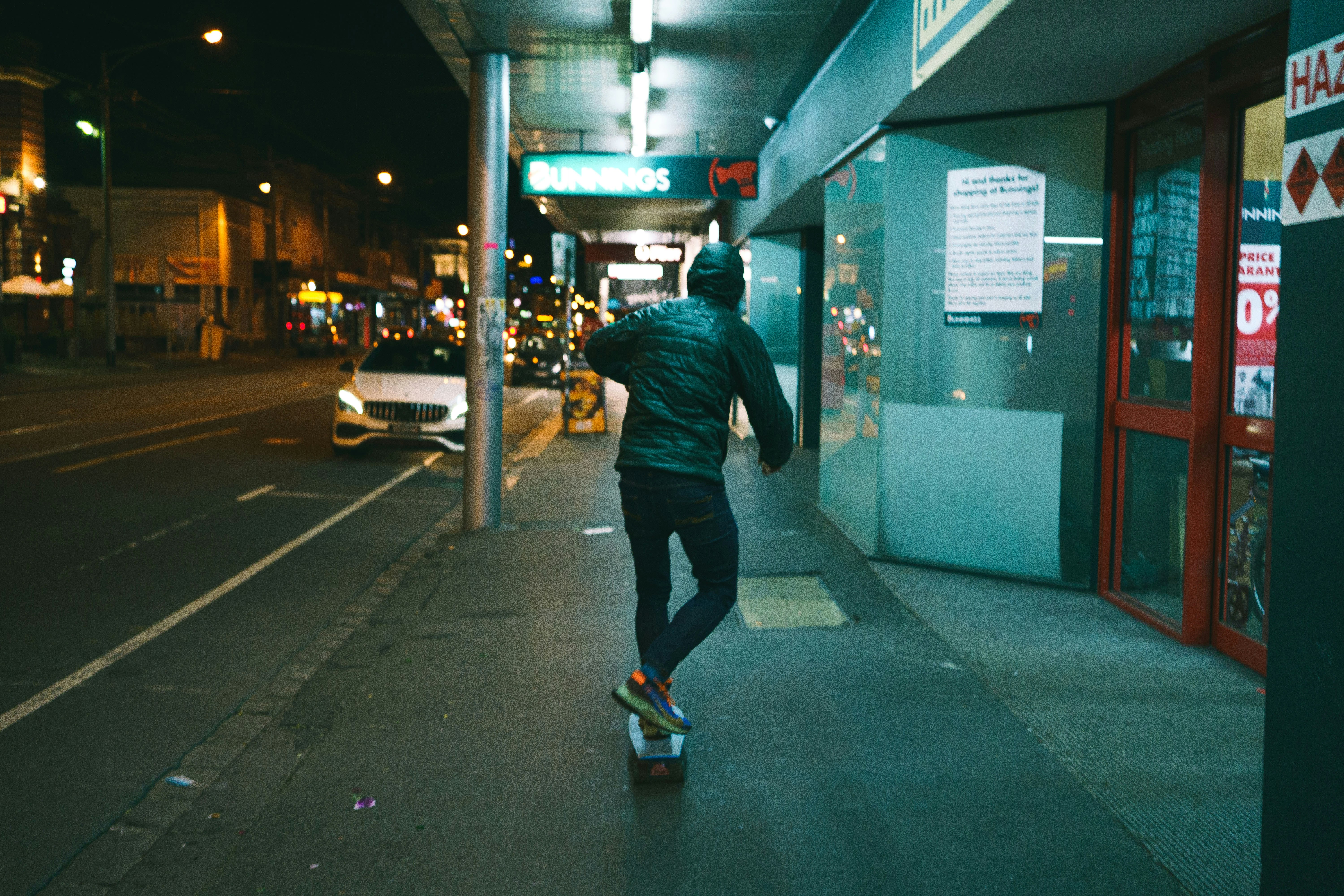 Person skateboarding along a dimly lit city sidewalk at night, surrounded by neon lights and storefronts.
