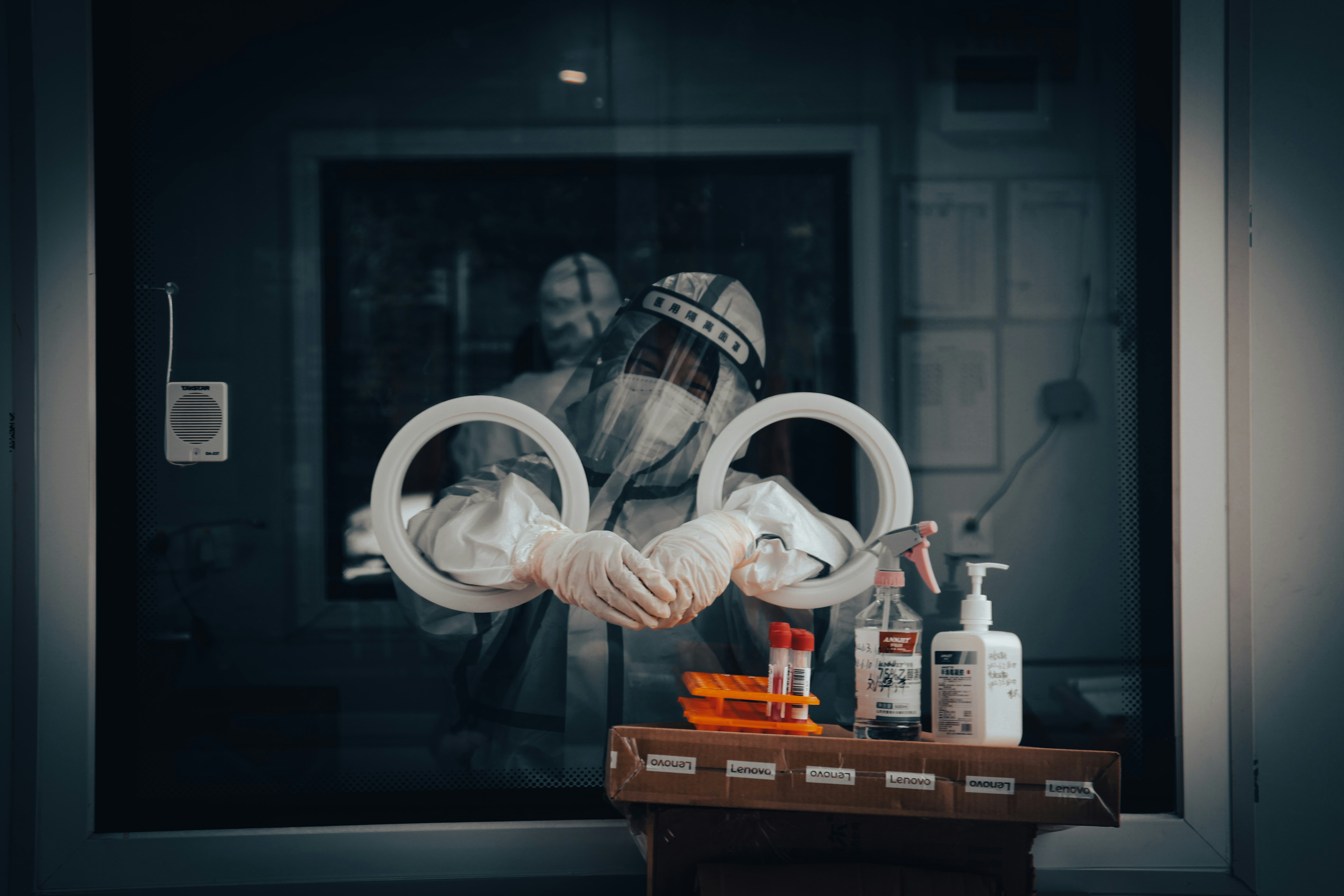 Healthcare worker in protective gear stands behind a glass barrier, surrounded by medical supplies. The image highlights the challenges of modern healthcare.