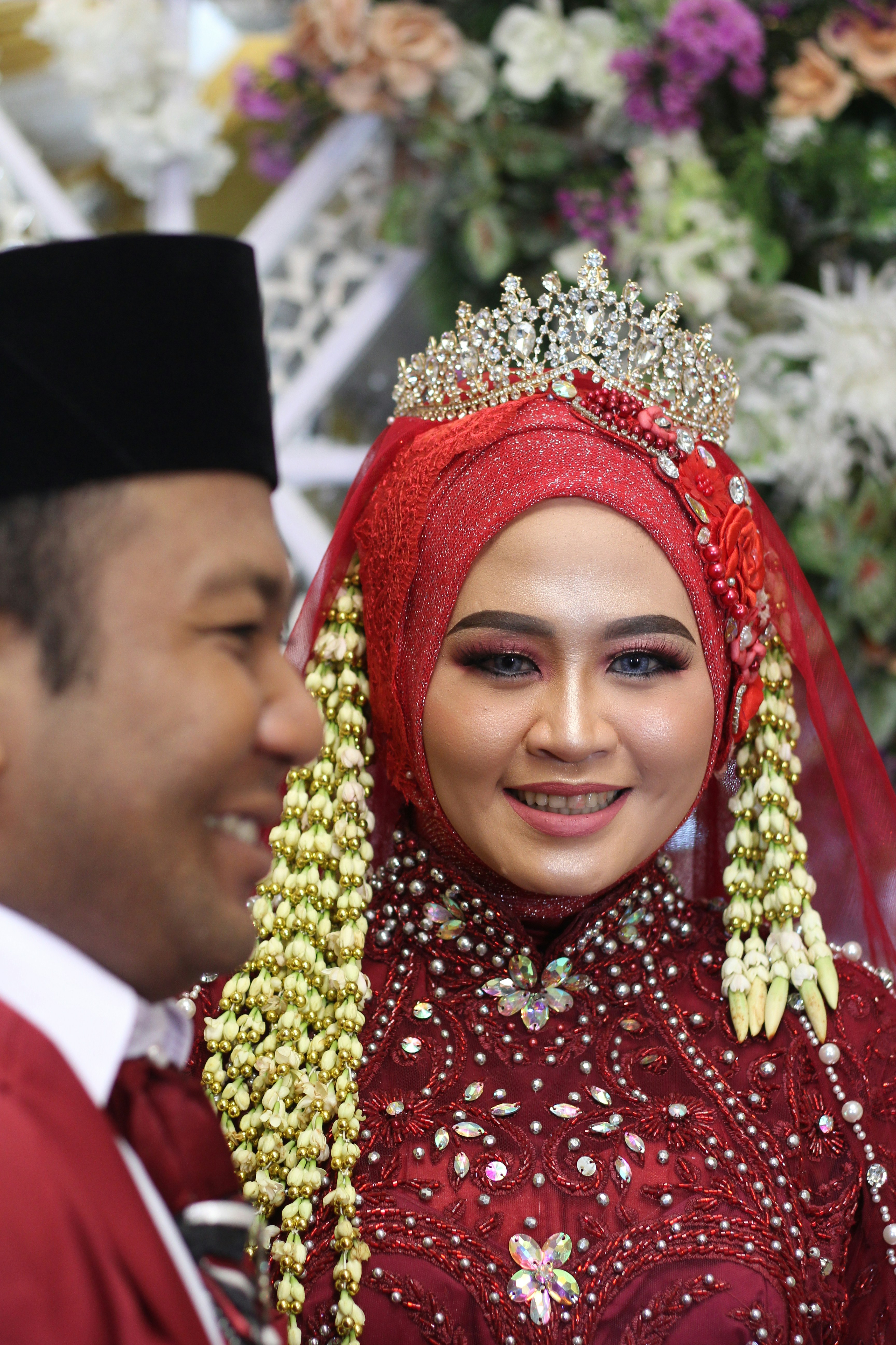 A bride adorned in a vibrant red gown and intricate jewelry smiles warmly, while a groom in traditional attire gazes at her. Floral decorations create a festive backdrop.