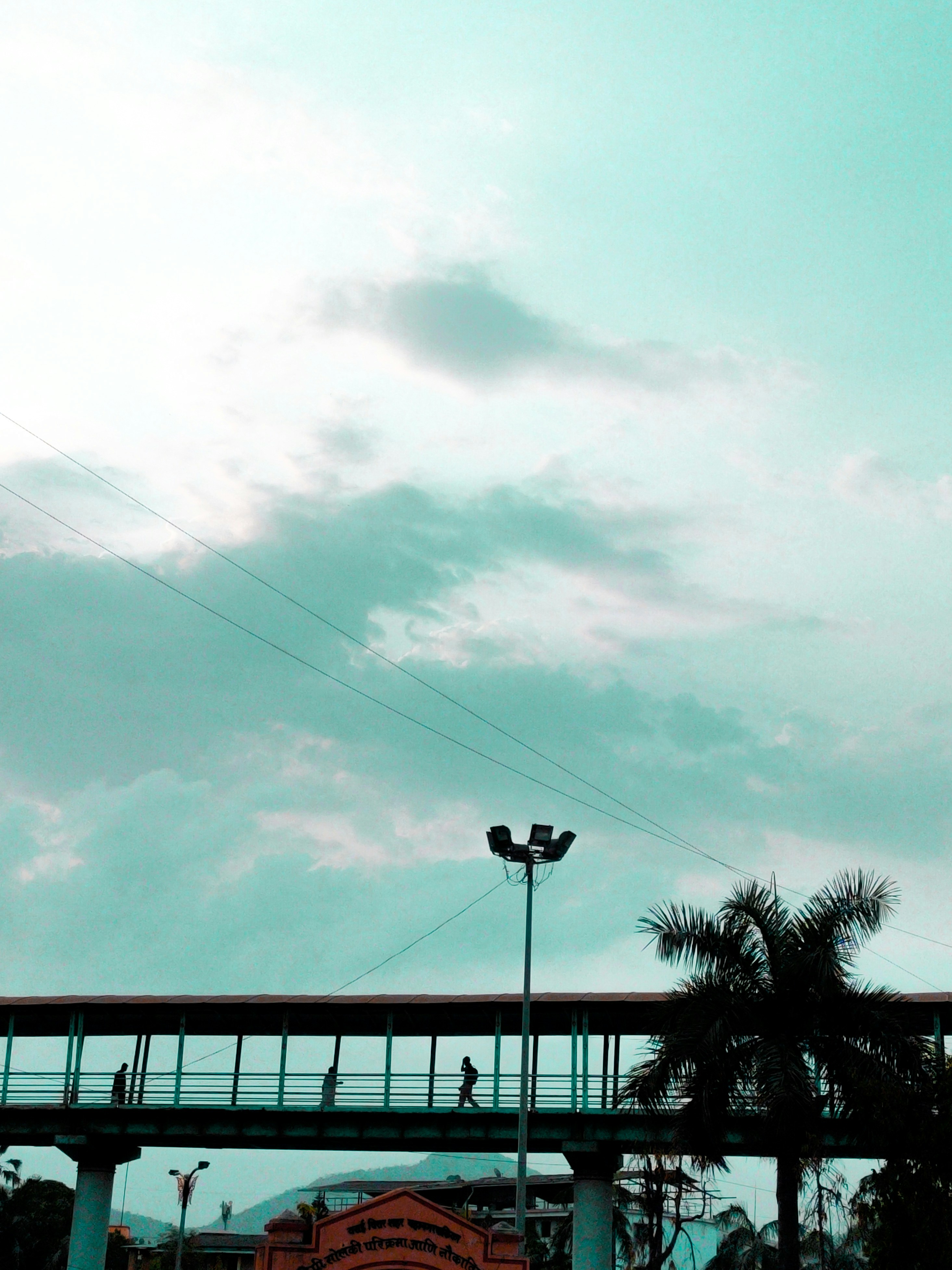 Silhouette of a person walking across a pedestrian bridge, surrounded by a moody sky and urban elements. The scene captures a moment of solitude in a bustling environment.