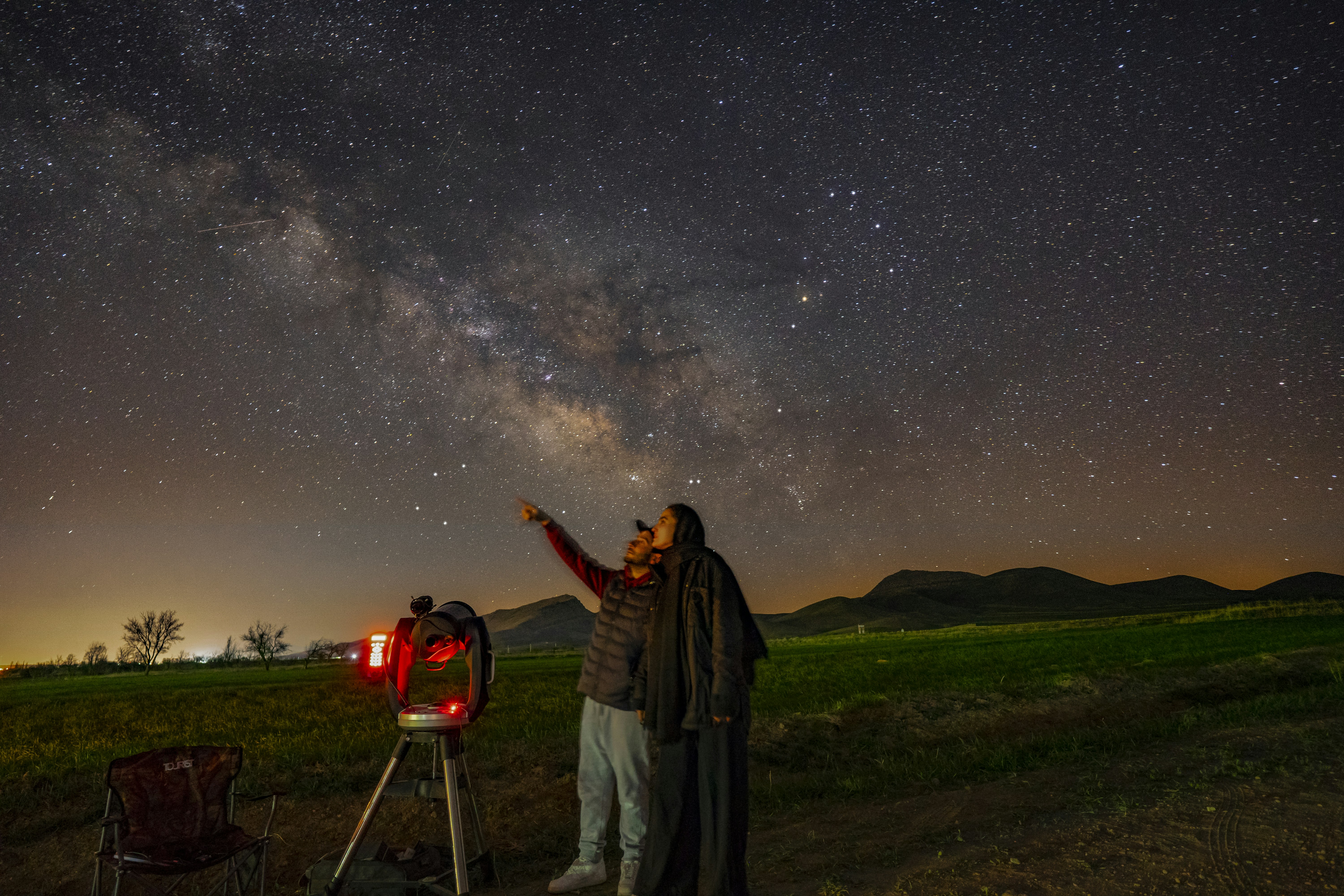 Two people stargazing with a telescope under a vibrant Milky Way sky in a rural landscape.