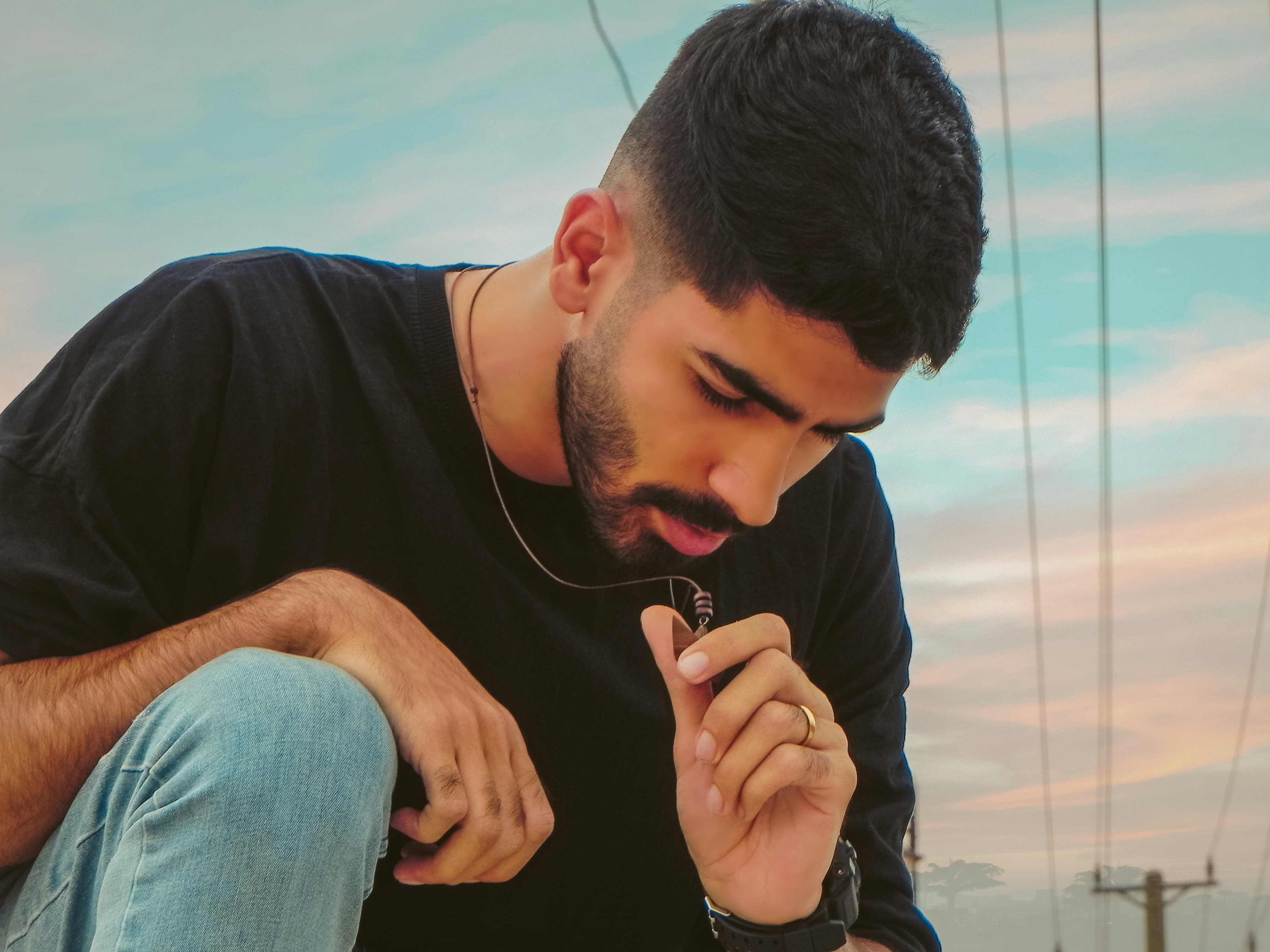 Young man in a black shirt thoughtfully gazing at a small object while seated outdoors, with a colorful sky and power lines in the background.