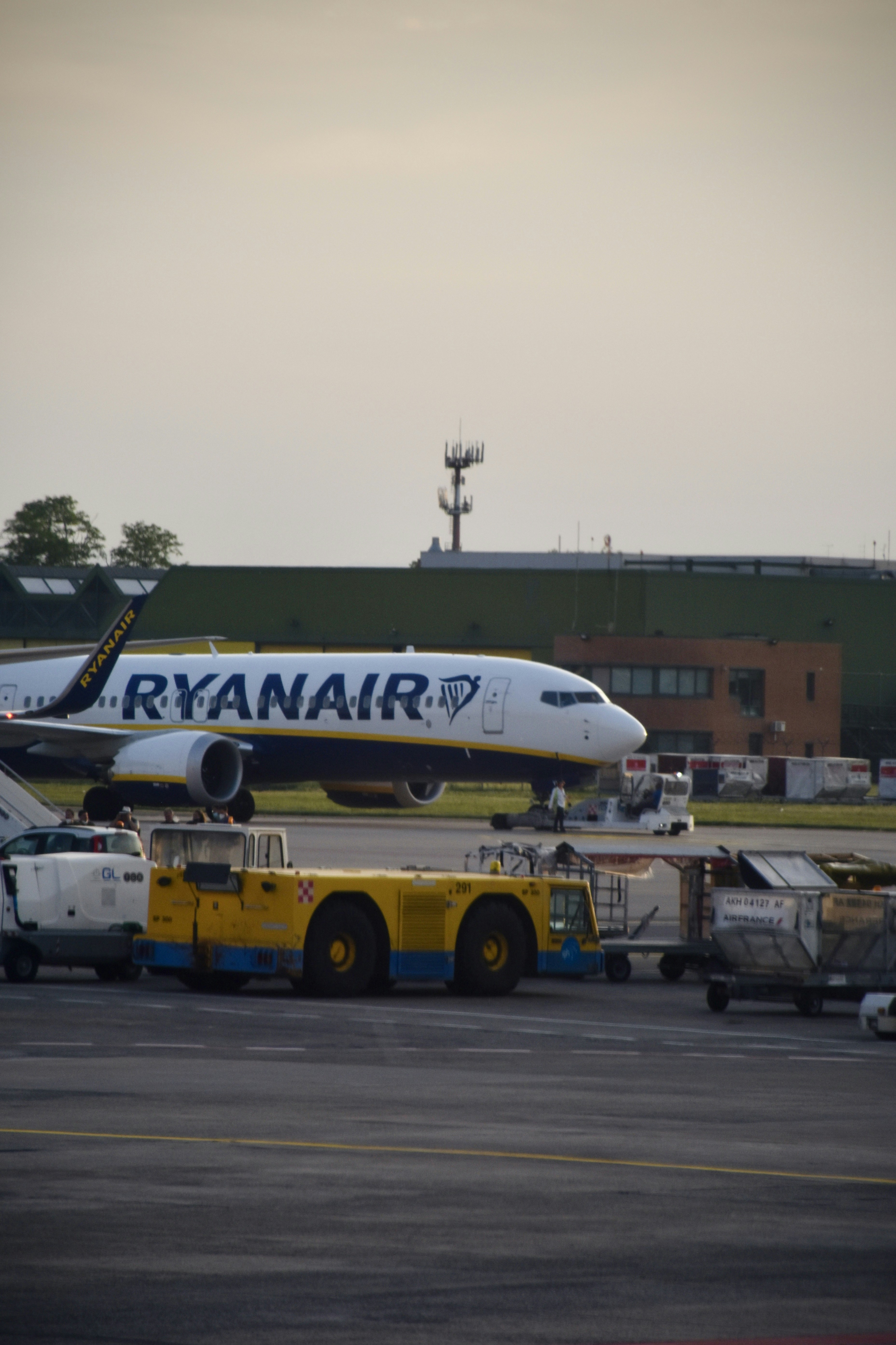 Ryanair aircraft taxiing on the runway with service vehicles and airport buildings in the background.