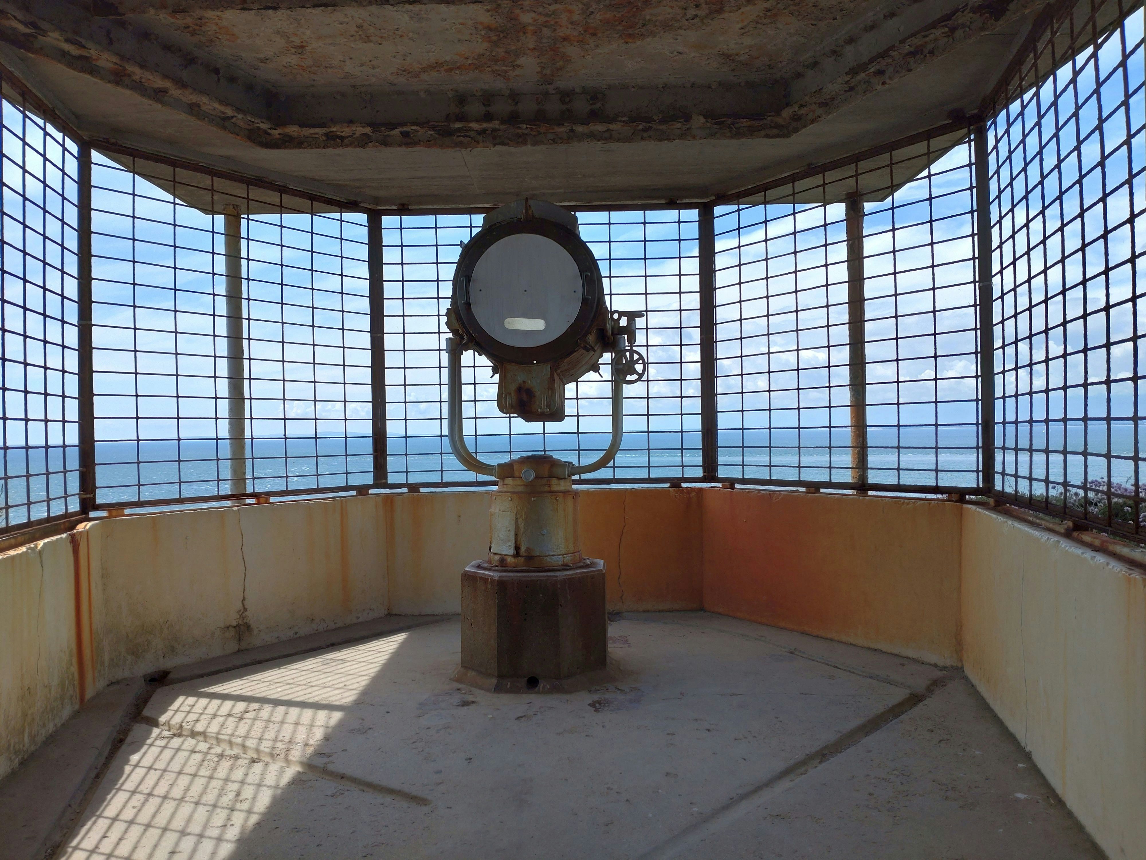 Interior view of a lighthouse lantern room featuring a large lens, surrounded by a grid of windows overlooking the sea.