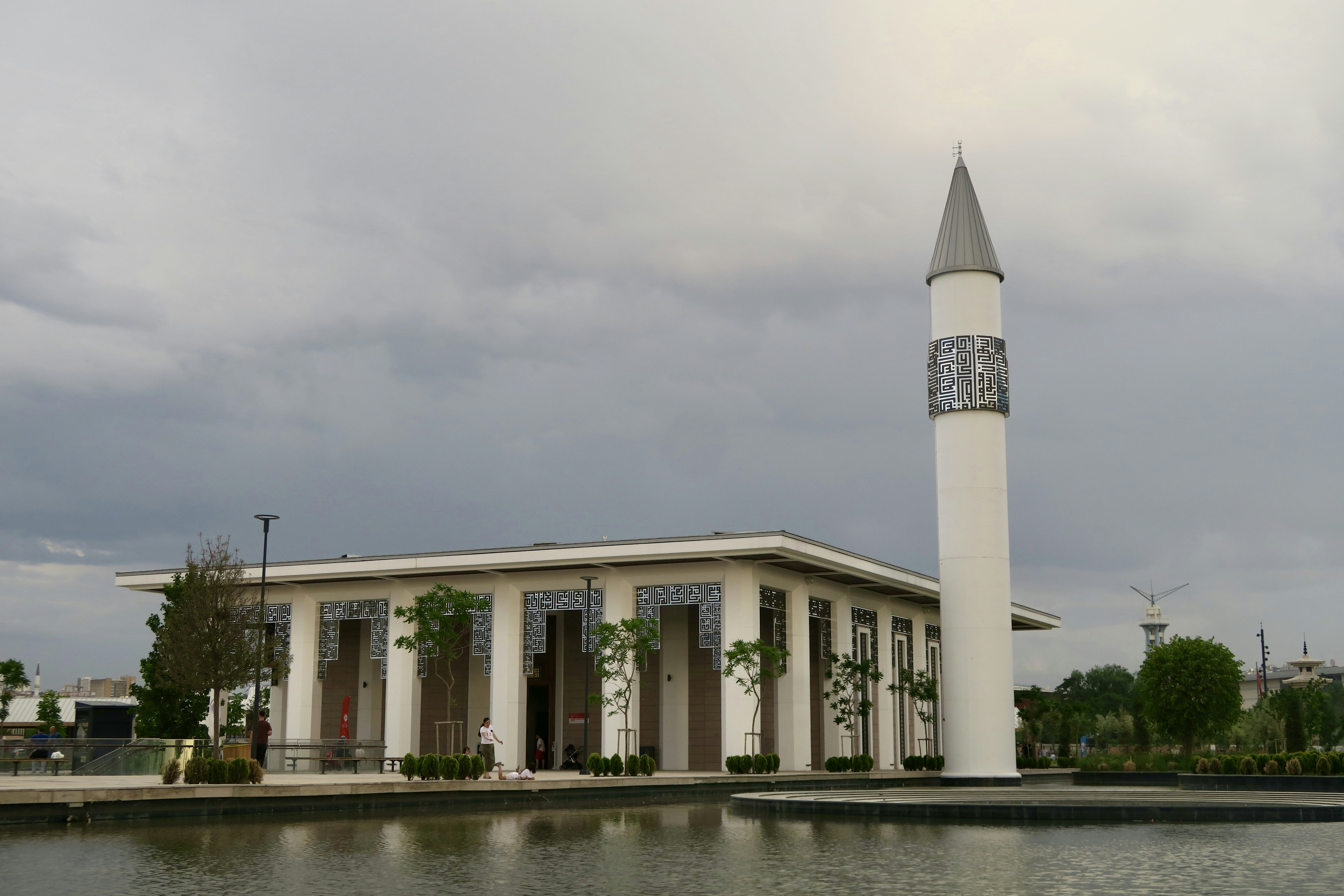 Contemporary mosque structure with a tall minaret, surrounded by a serene water feature and lush greenery.