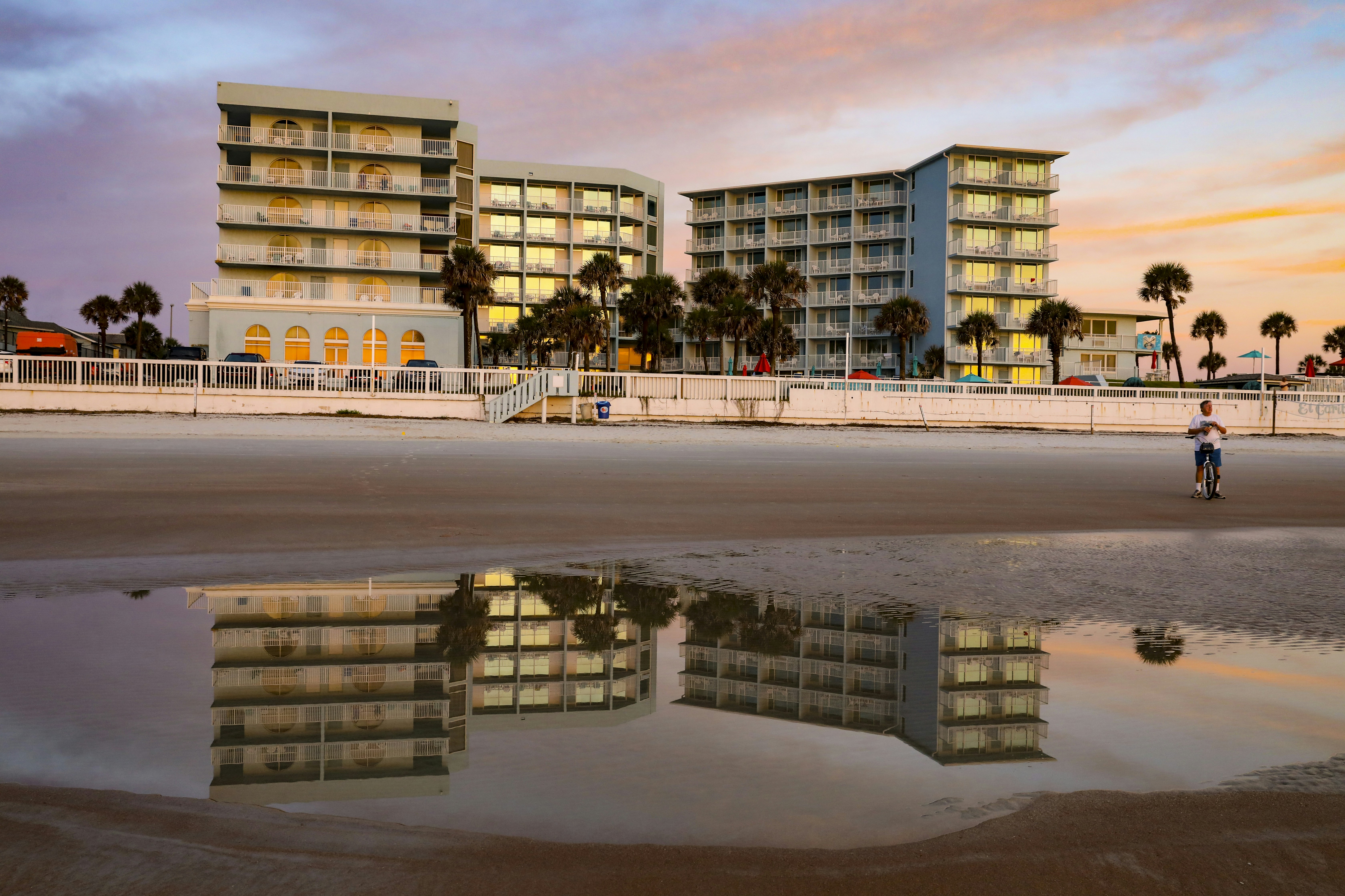 Coastal buildings reflecting in wet sand at sunset, with a cyclist in the foreground. The scene captures the tranquil essence of beach life.