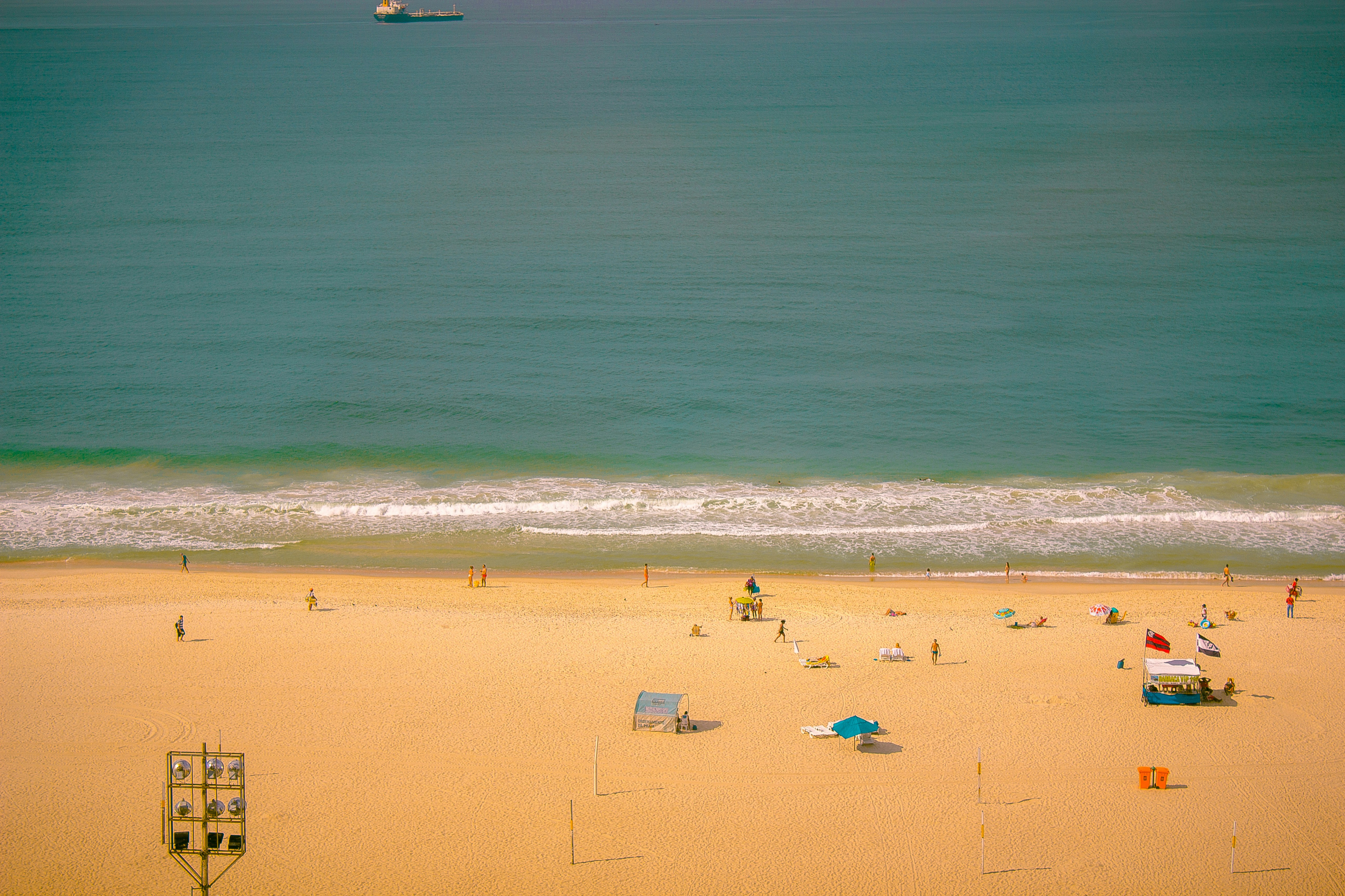 Vibrant beach scene with sunbathers and colorful umbrellas, framed by gentle waves and a distant ship on the horizon.