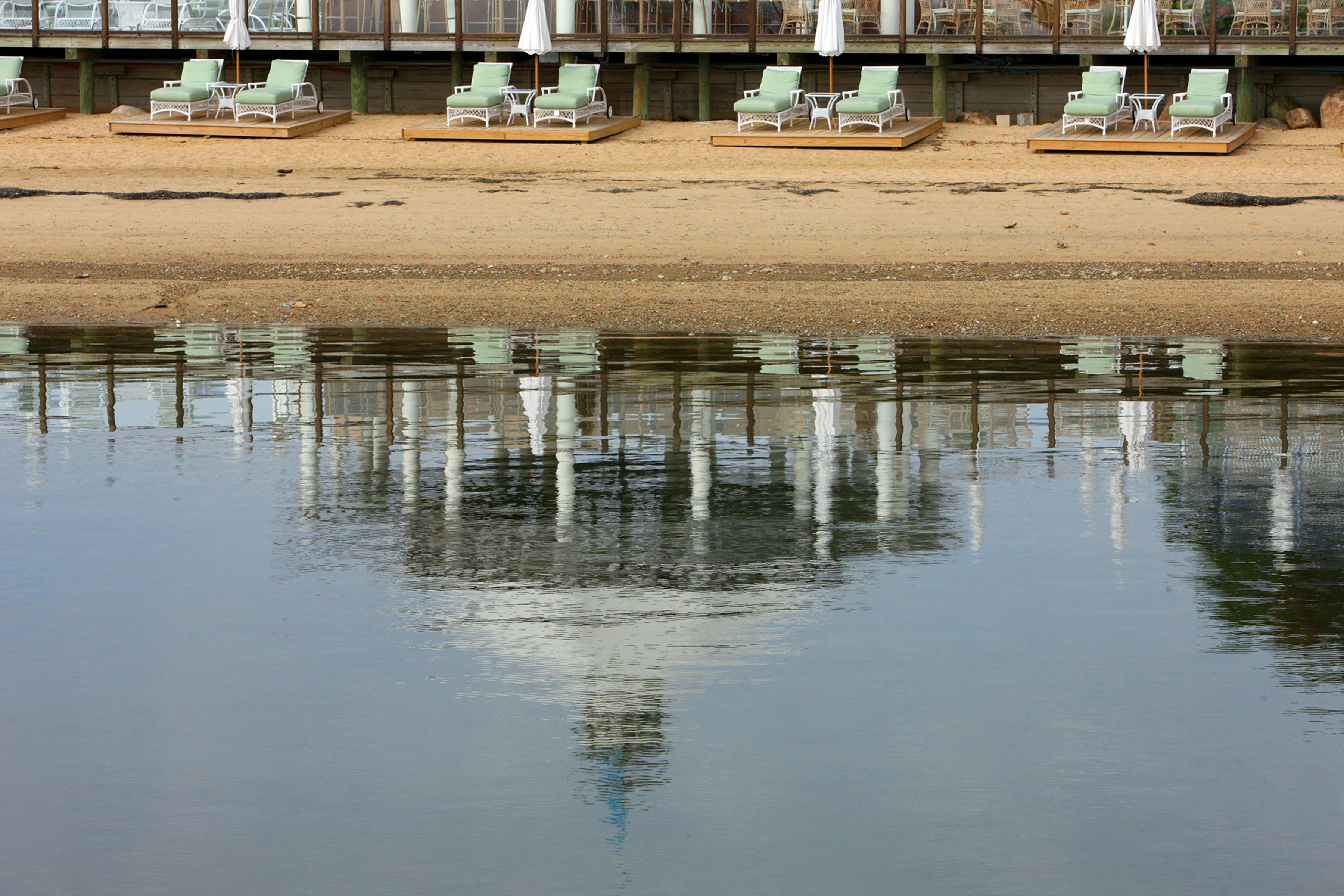 Elegant lounge chairs line the sandy beach, mirrored perfectly in the calm water below. The serene atmosphere invites relaxation.