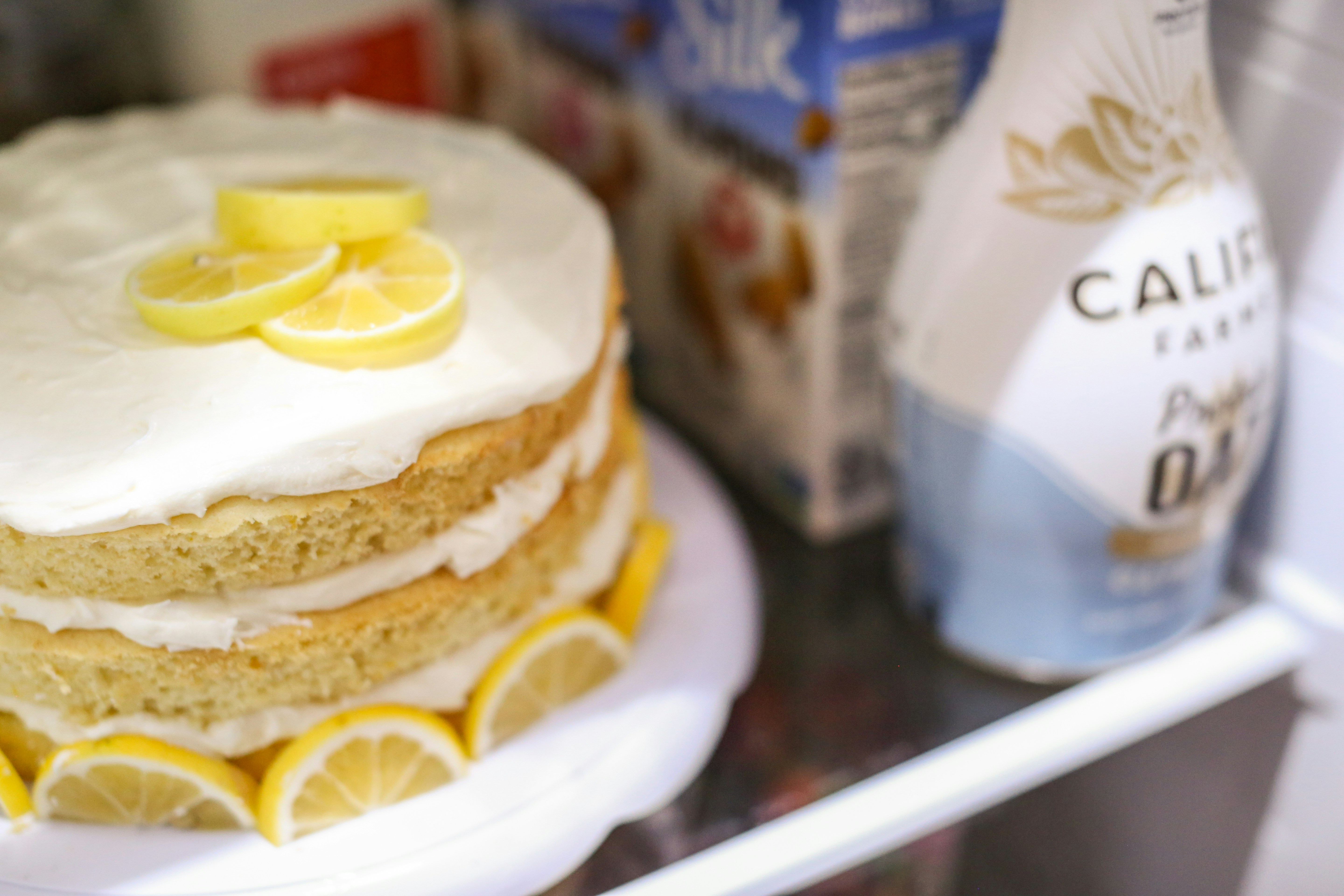 Lemon cake adorned with fresh lemon slices, nestled in a refrigerator surrounded by various food items. The cake's creamy frosting and vibrant decorations stand out.