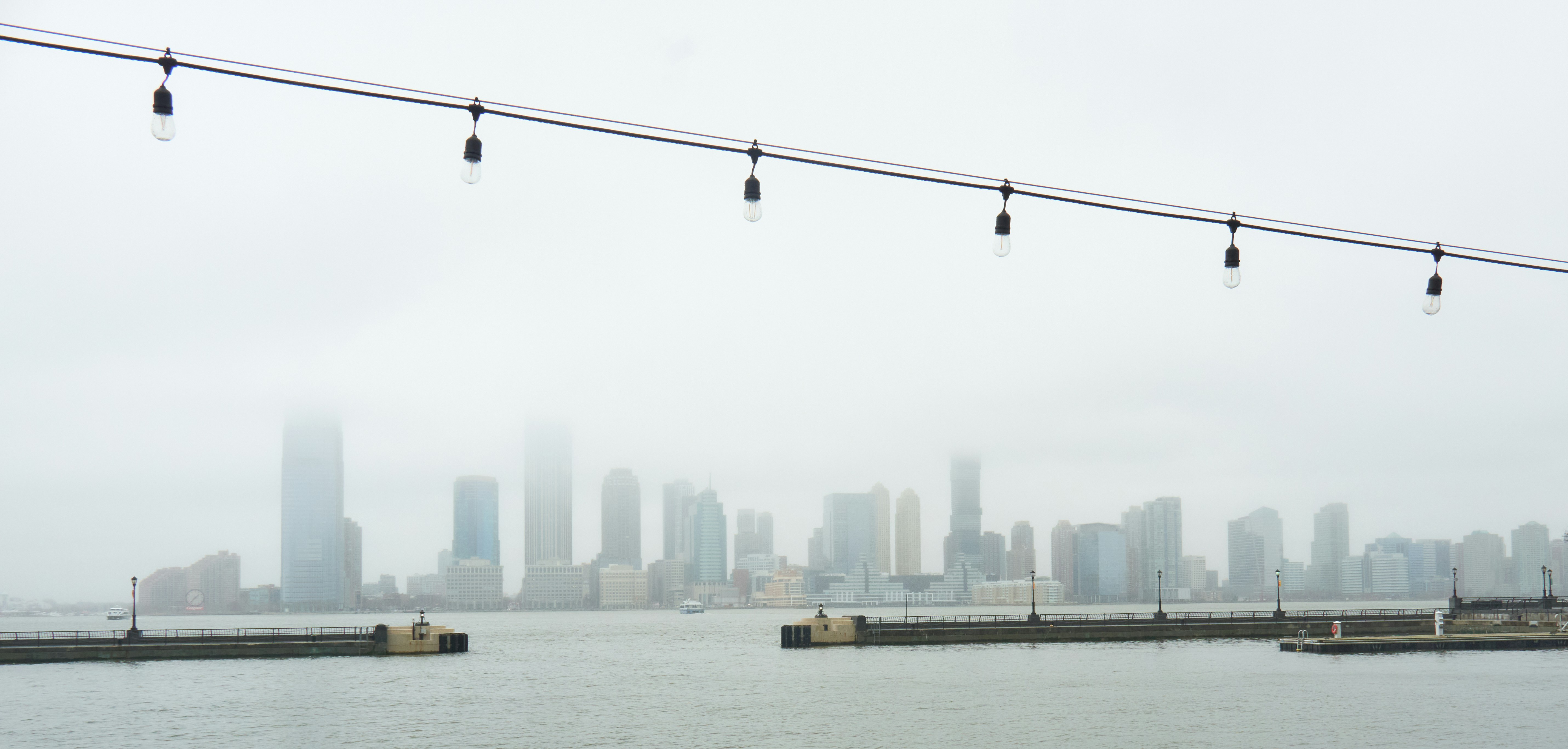 City skyline shrouded in mist across a tranquil body of water with overhead string lights.