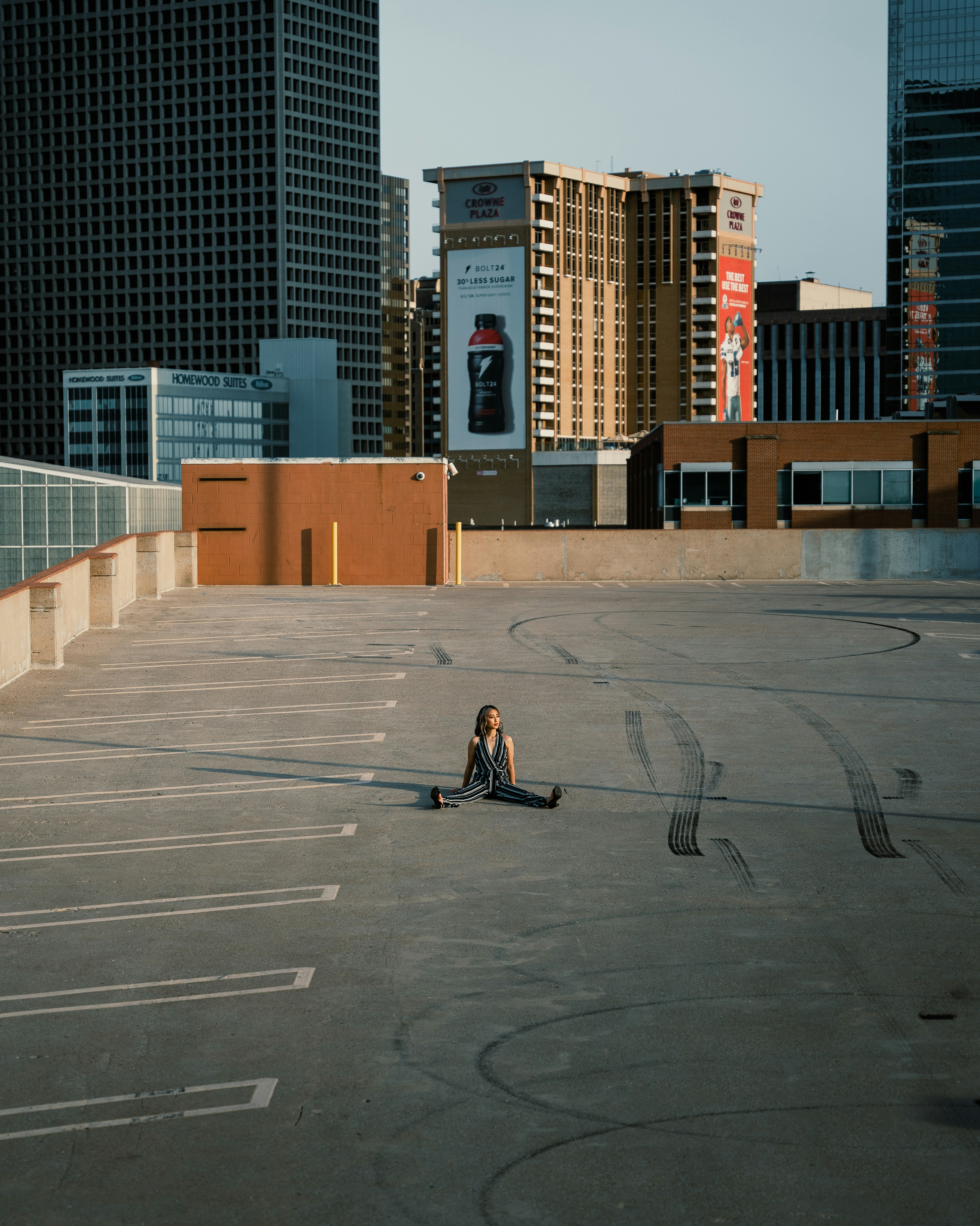 Child seated in a striped outfit on an empty rooftop parking lot, surrounded by towering city buildings and distant billboards.